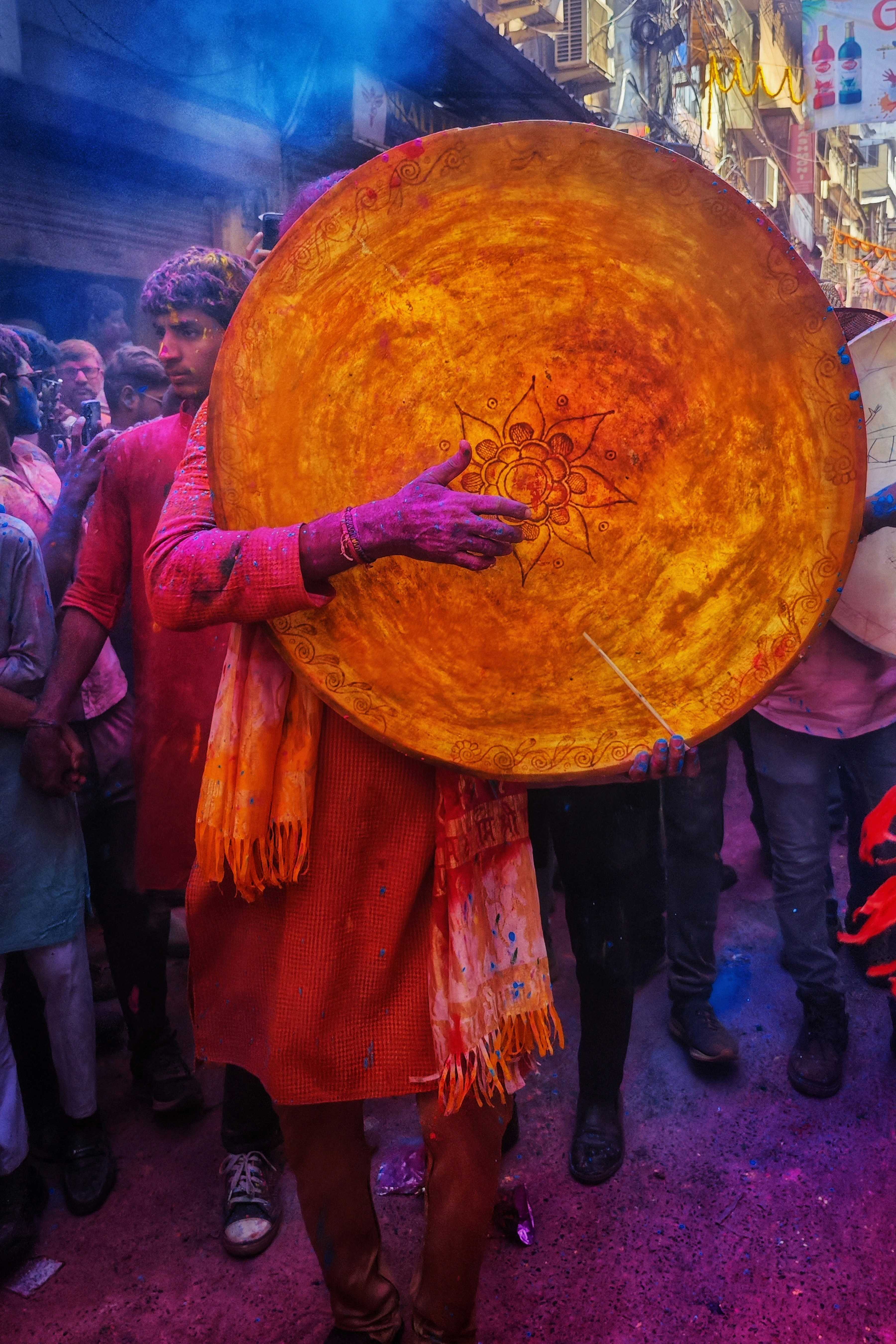 A vibrant celebration scene featuring a person holding a large, colorful drum, surrounded by a crowd adorned in festive attire. The atmosphere is filled with joy and color.