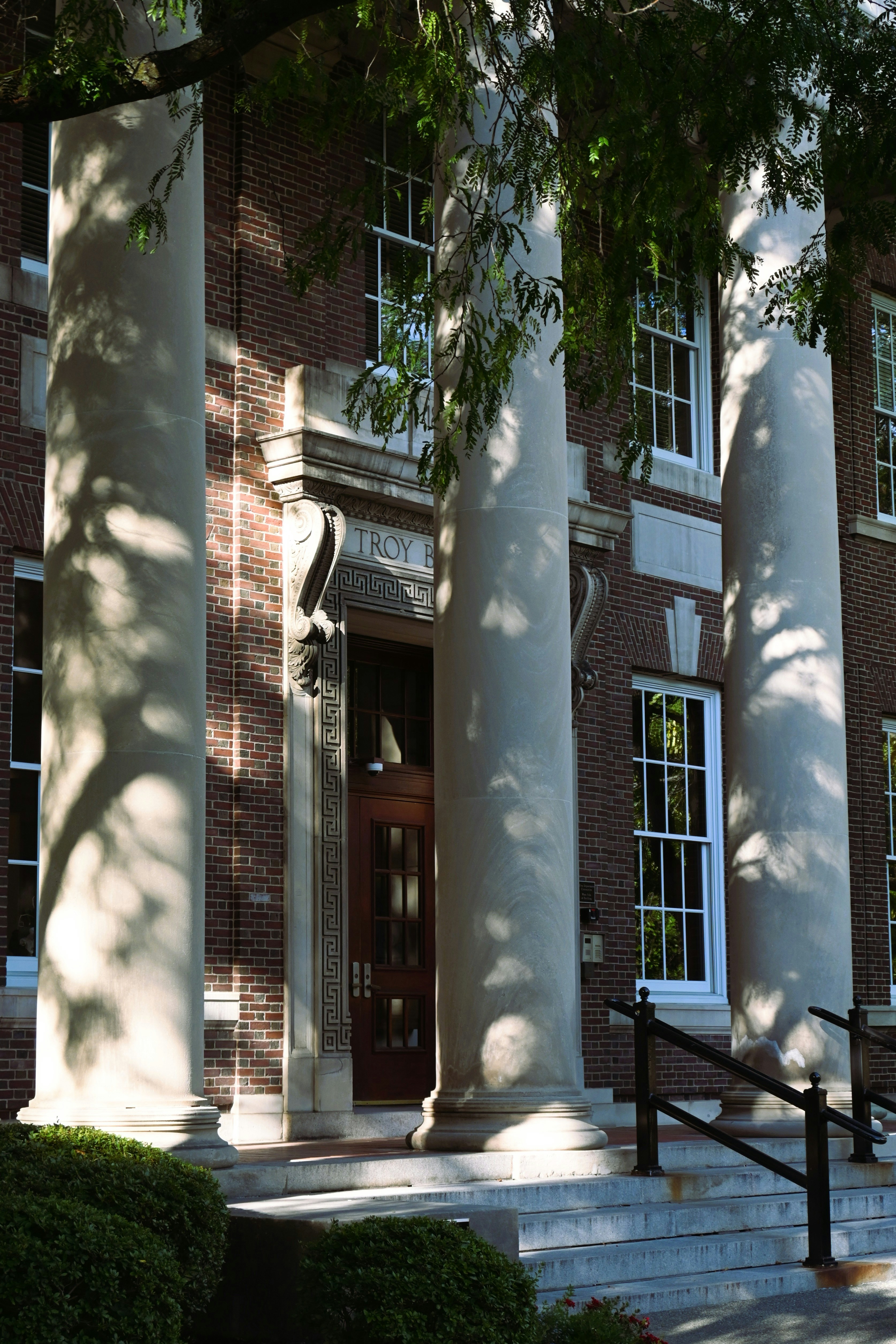 Historic entrance of the Troy building framed by grand columns and dappled sunlight filtering through the trees.