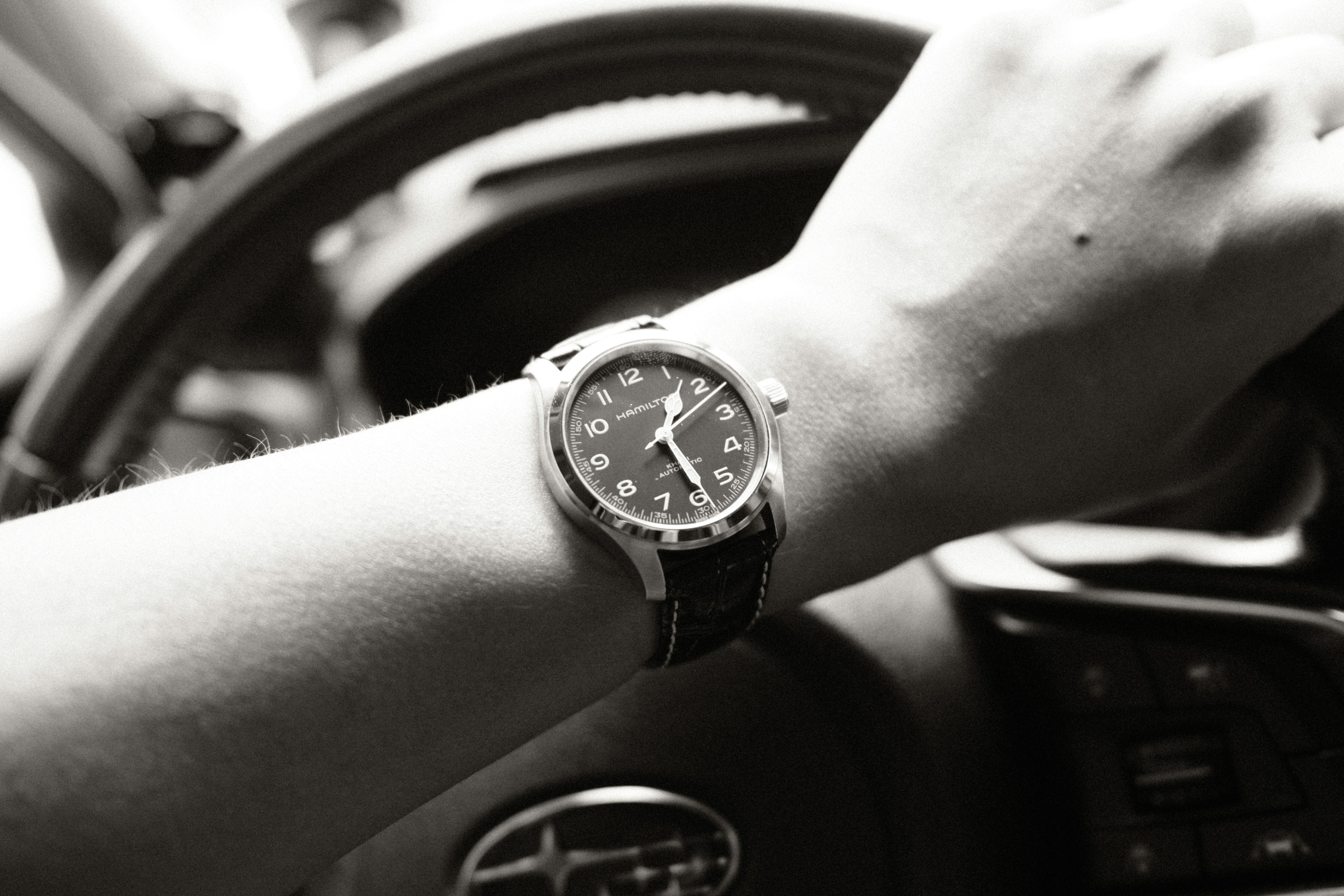 A hand adorned with a classic watch rests on a steering wheel, emphasizing the blend of timekeeping and driving experience.