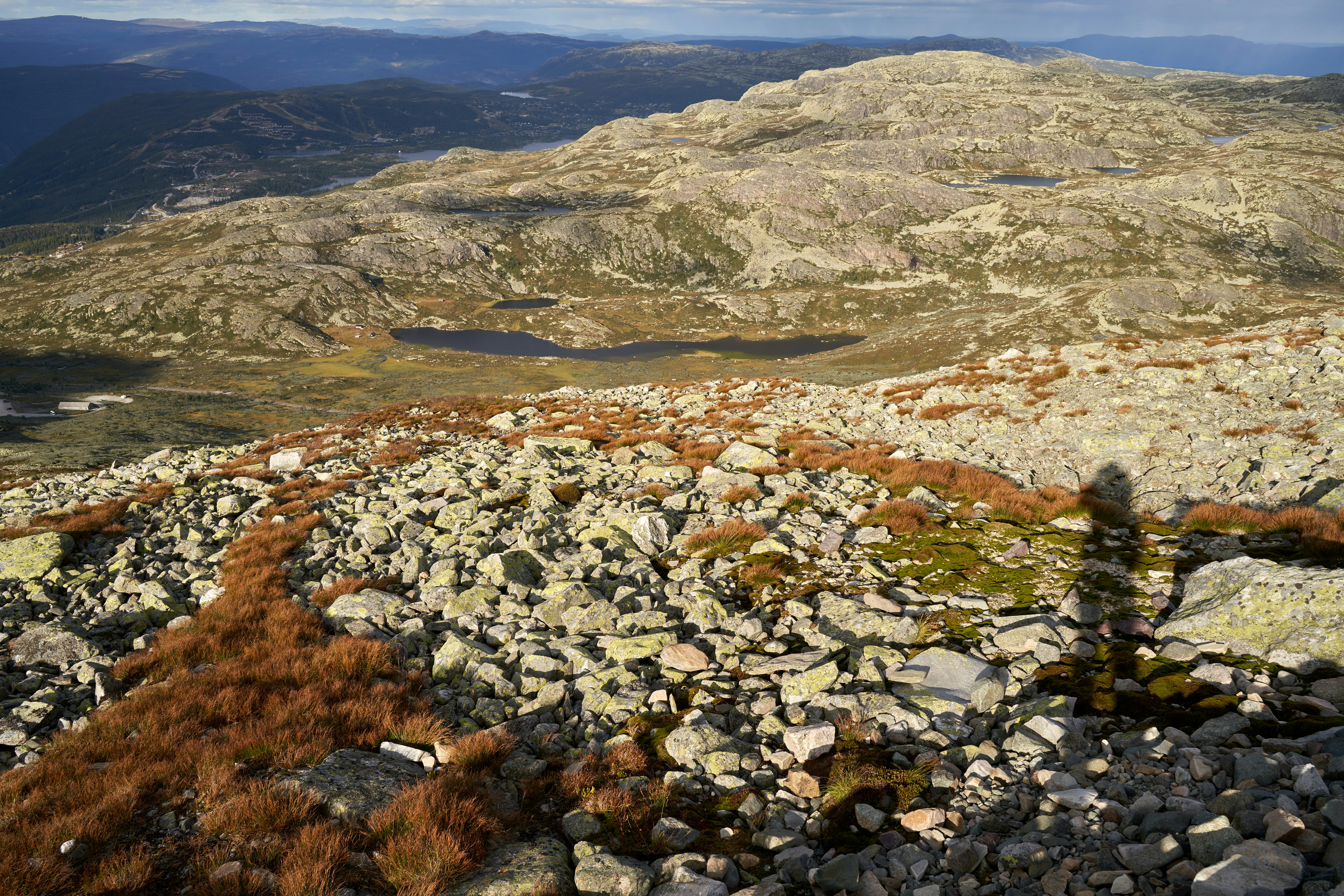 Majestic View from Gaustatoppen, Telemark's highest mountain at 1883 meters, offers a breathtaking panorama of colorful, rocky peaks, lush grasslands, and dense pine forests and the photographer's shadow | Rocky mountain landscape with small lakes under sky