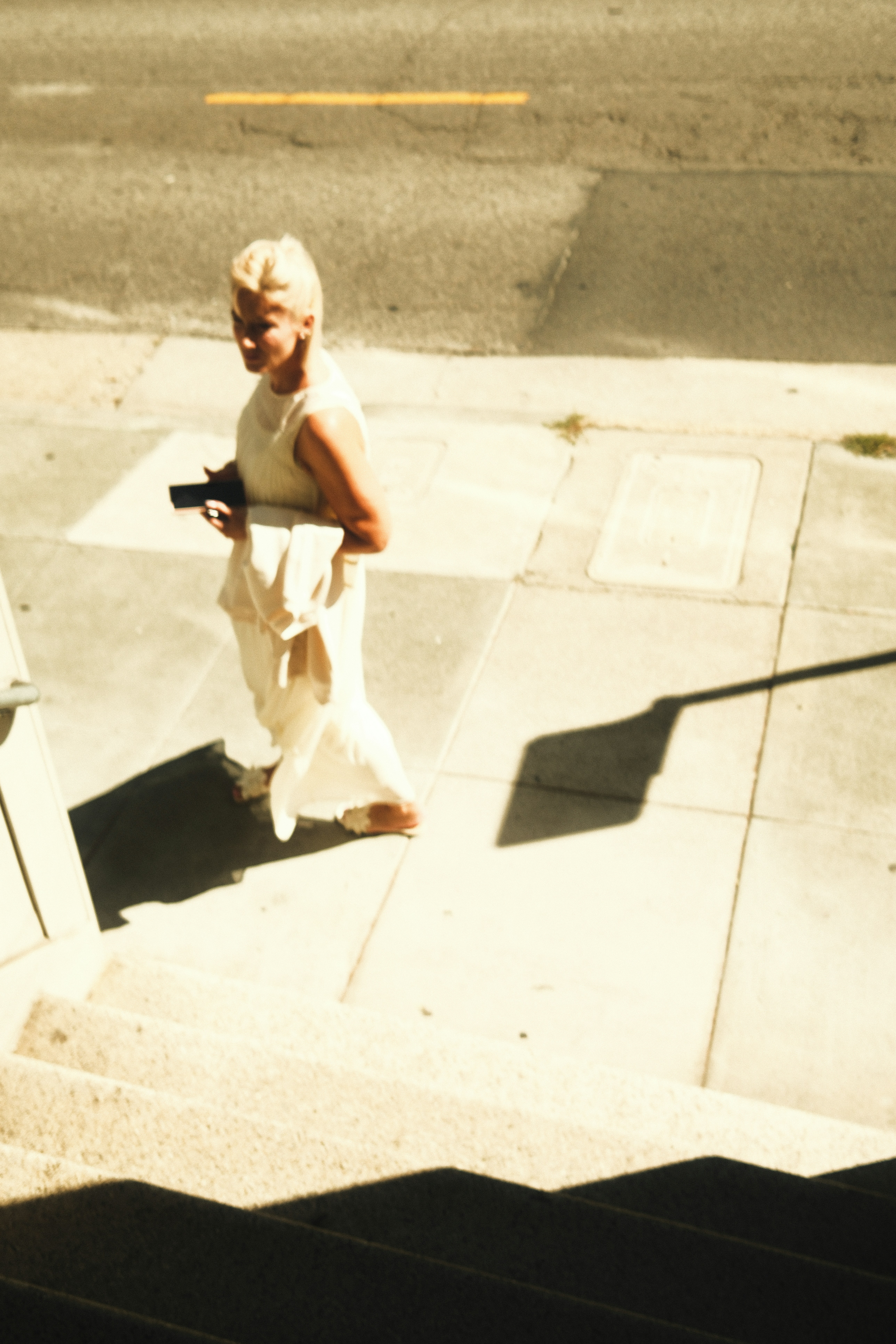 A woman in a flowing white dress walks down the steps, holding a phone and a light shawl, with shadows cast on the pavement. The urban setting adds a contemporary touch.