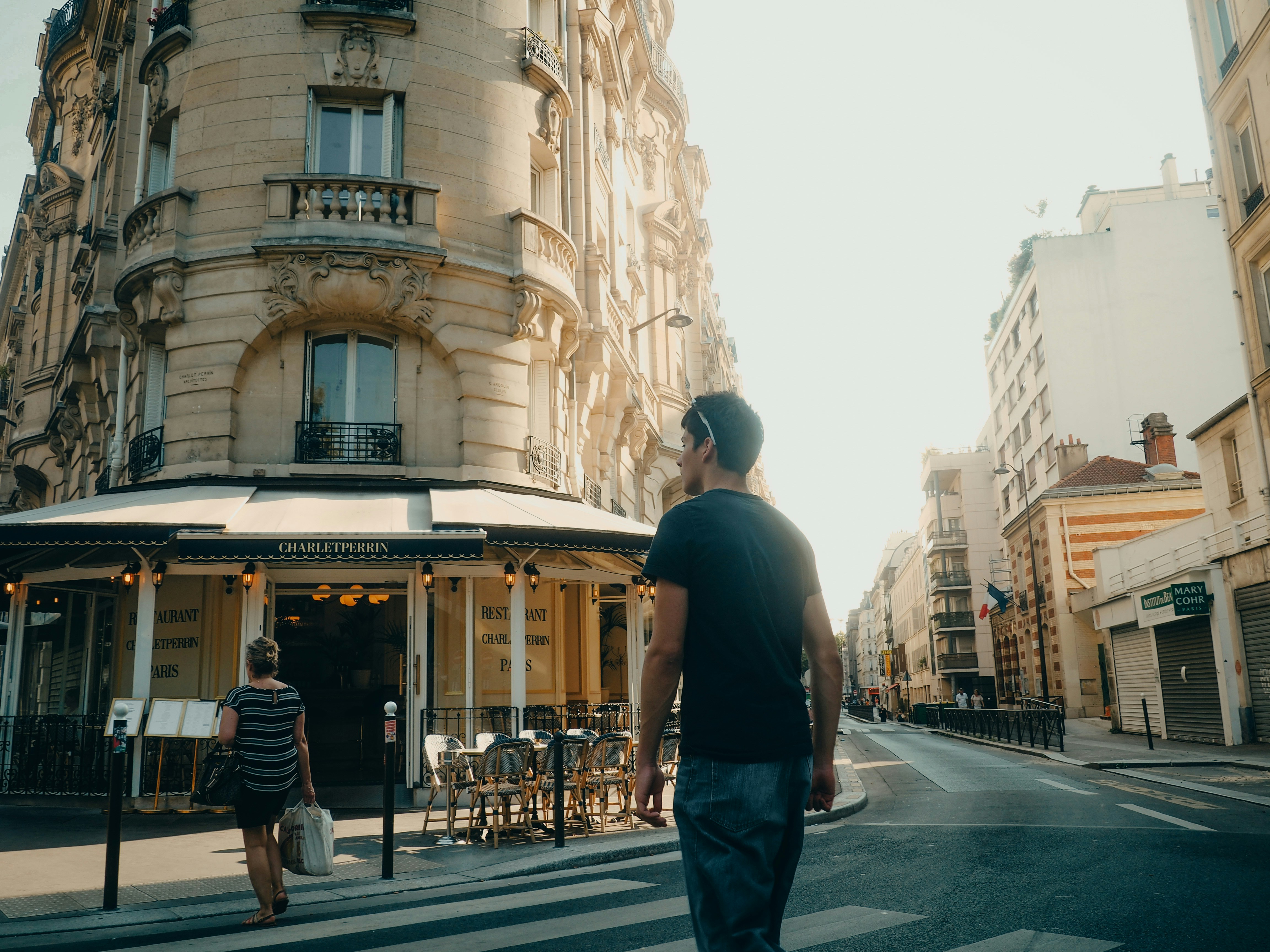 This photo captures the essence of Paris street photography: elegant Haussmann architecture, golden facades, classic French windows, ornate balconies, and stylish urban design. The scene shows two people walking, blending daily life with the historic atmosphere of the city. With cars, street lamps, and shadows, this is pure urban lifestyle in the heart of Paris, France. The perspective lines highlight the beauty of Parisian architecture, while the casual street scene adds authenticity. Perfect for travel photography, cityscape lovers, and anyone searching for the timeless charm of the French c | Man walks down a sunny european street.