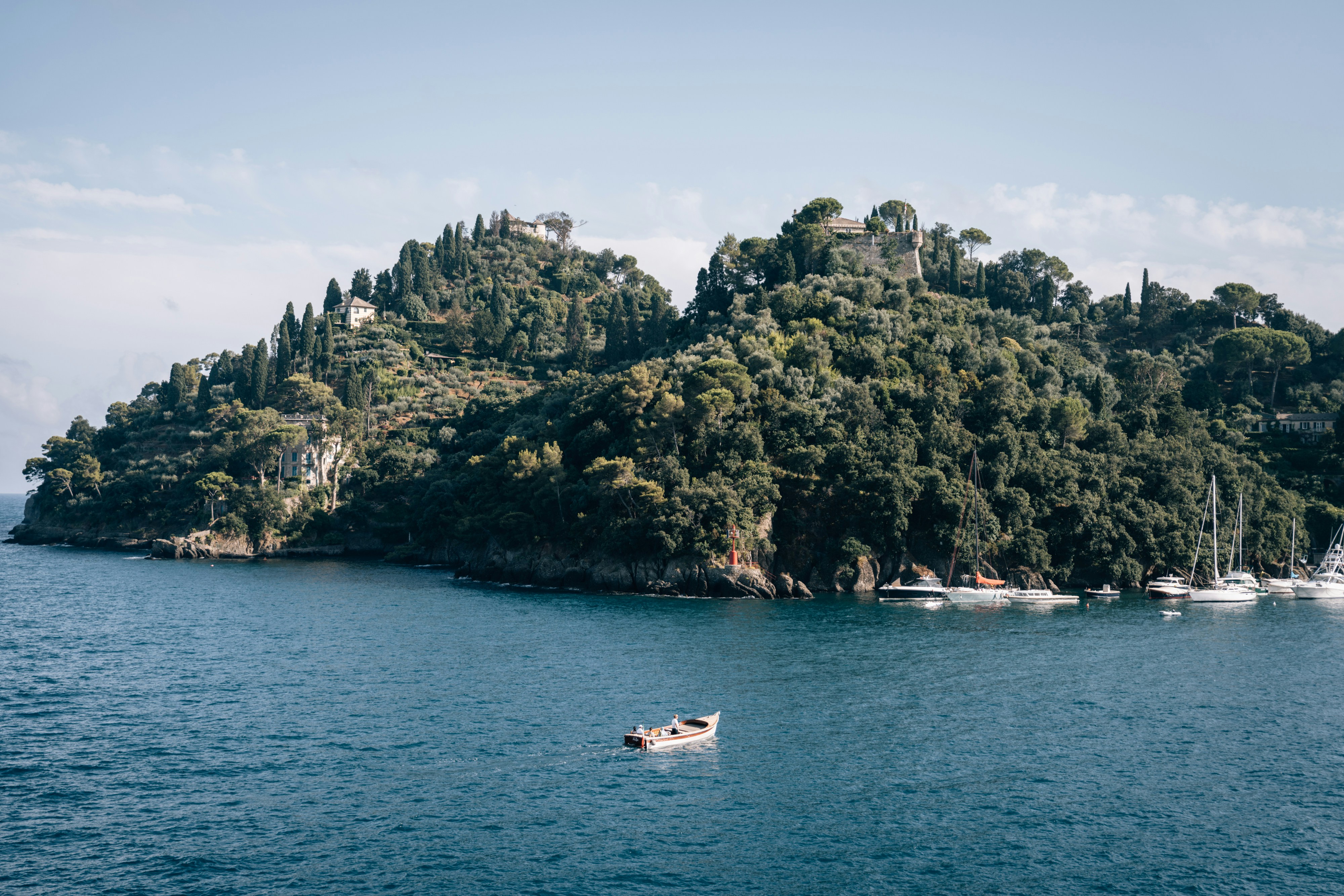 A small boat glides through tranquil waters near a lush, green island dotted with elegant villas and sailboats. The scene captures the essence of coastal relaxation.