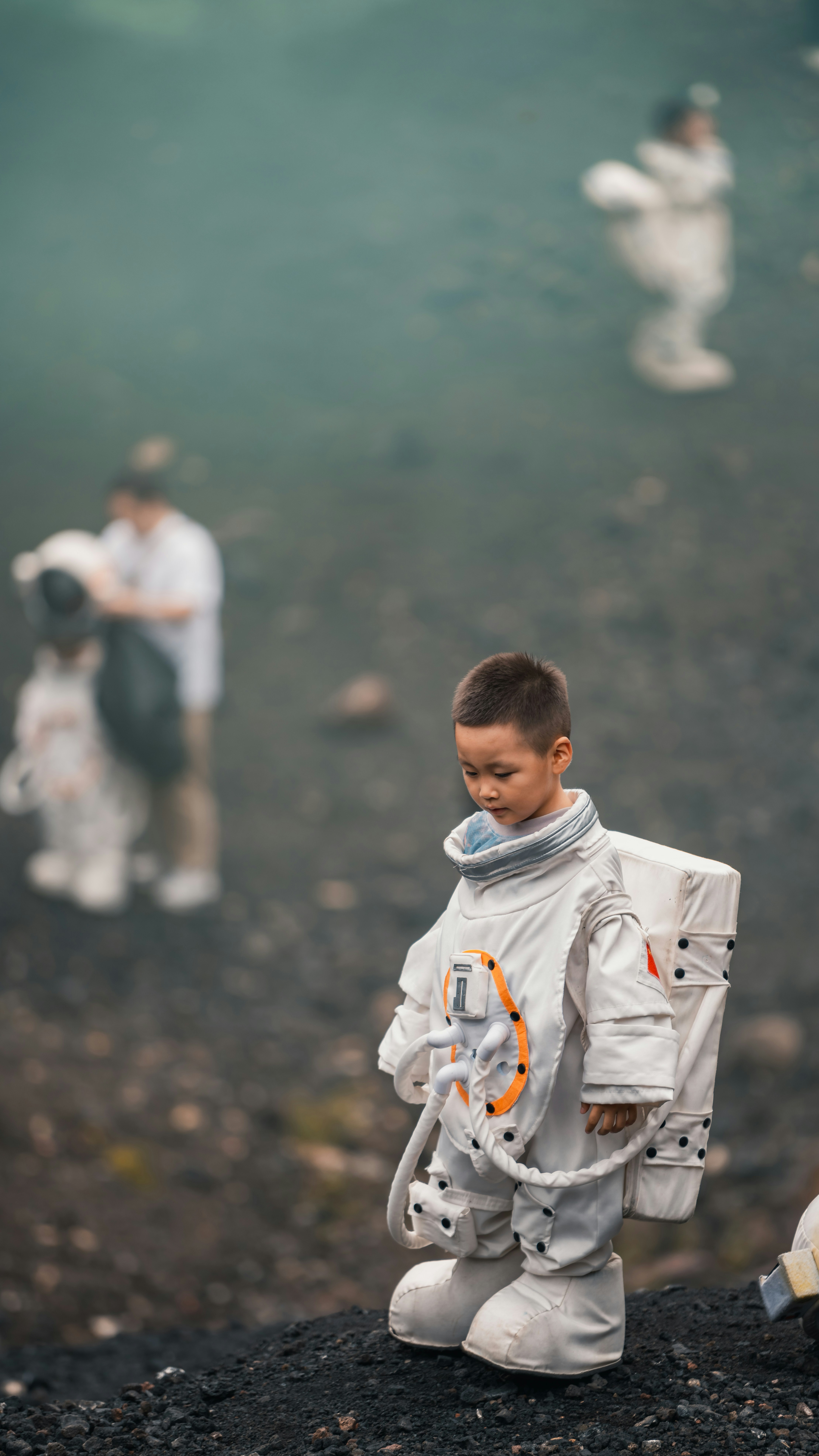 Young child dressed in a space suit stands on rocky terrain, gazing down thoughtfully, while other figures are blurred in the background.