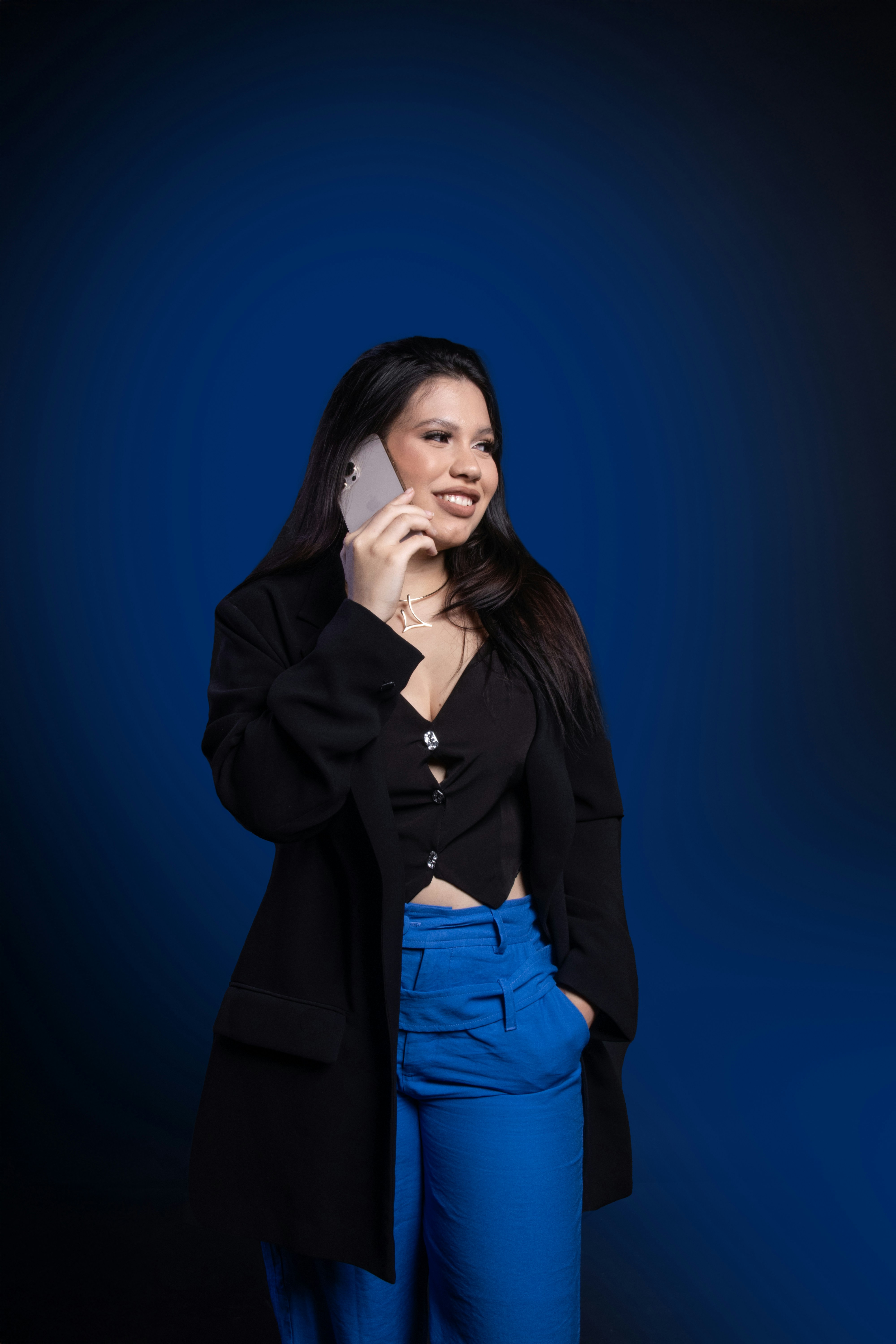 Young woman in a black blazer and blue pants, engaged in a phone conversation against a deep blue background.