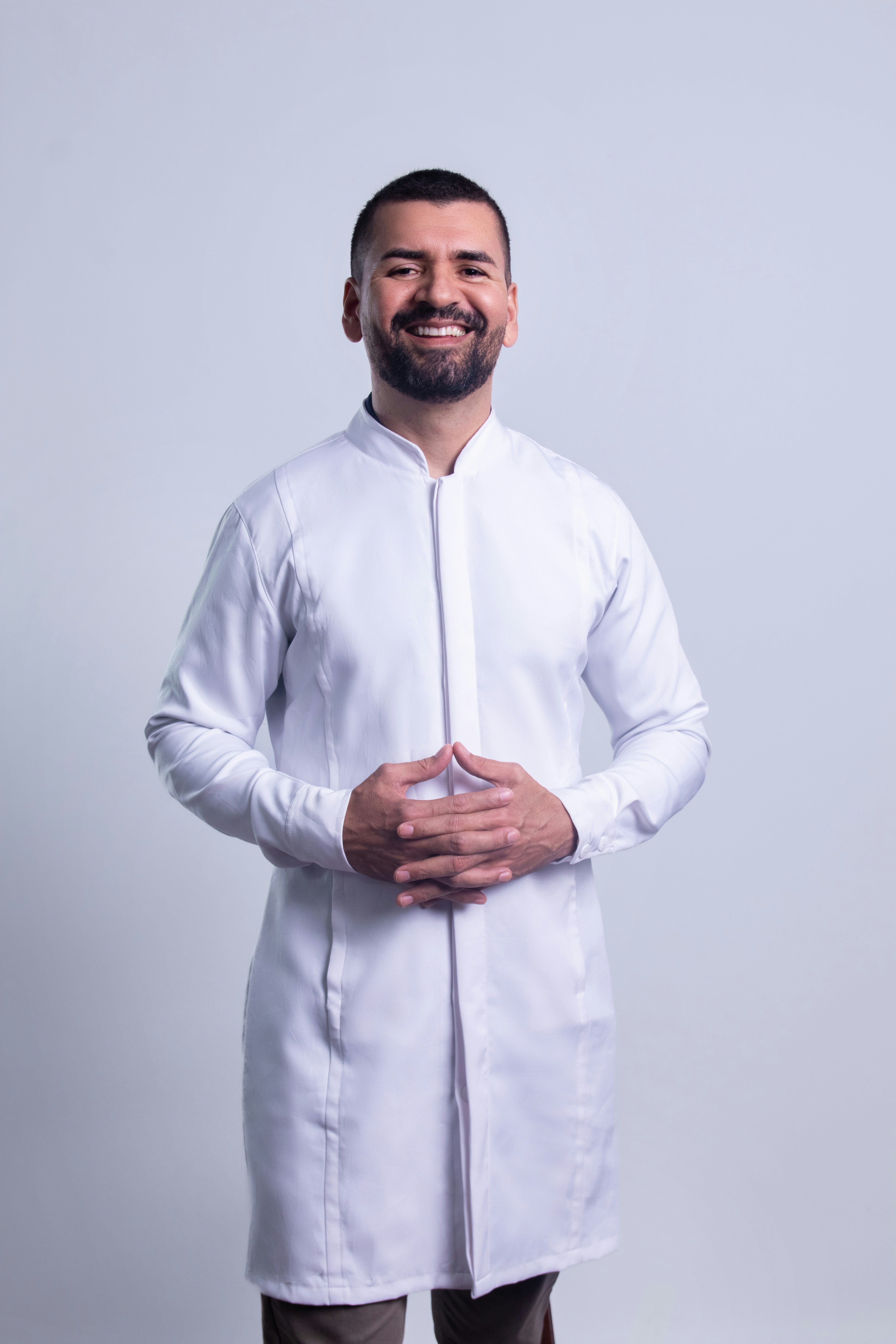 Smiling man in a white coat poses confidently with hands clasped, set against a light gray background.
