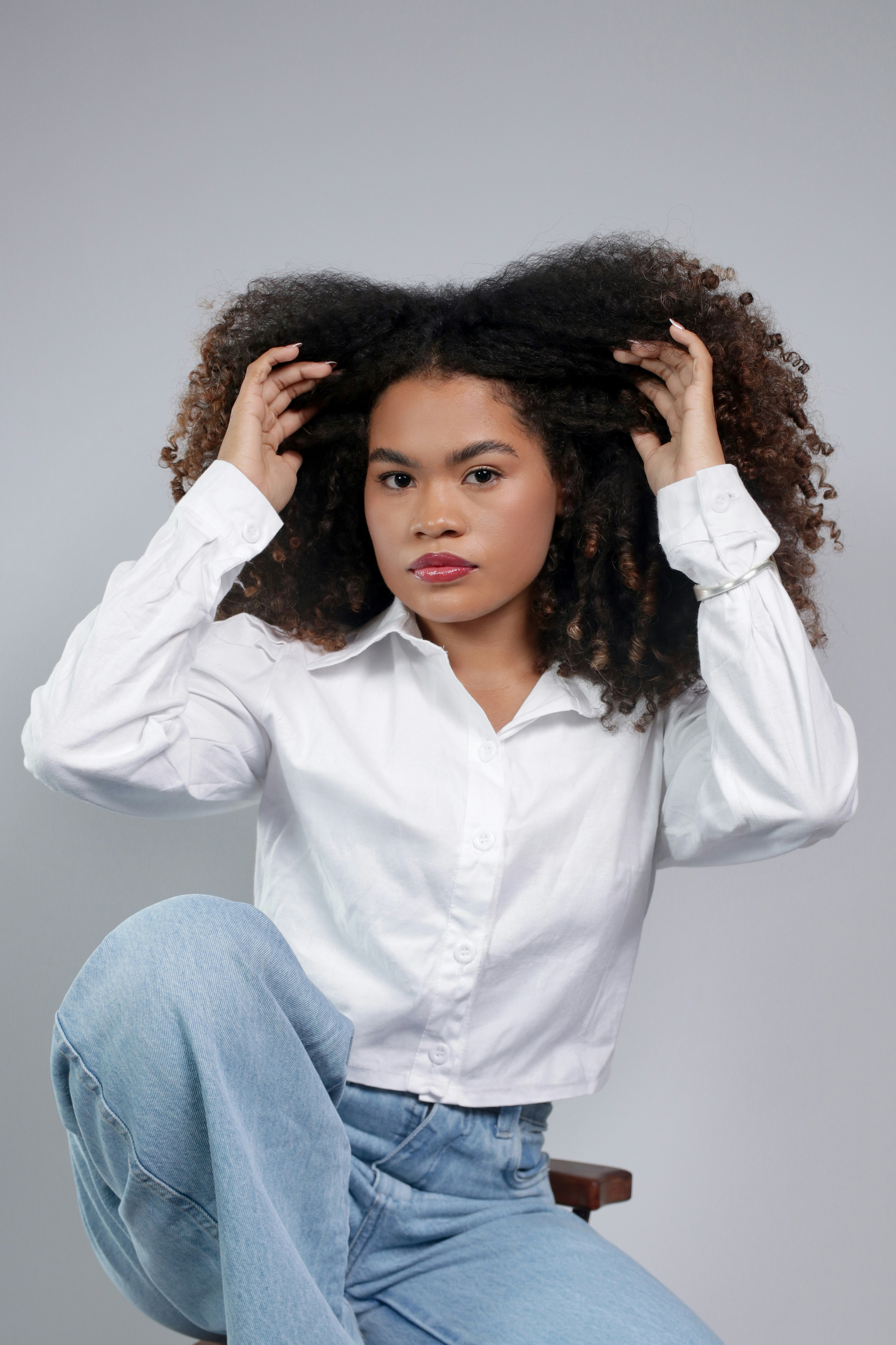 Young woman with voluminous curly hair styled dramatically, wearing a white shirt and blue jeans, striking a confident pose against a neutral background.