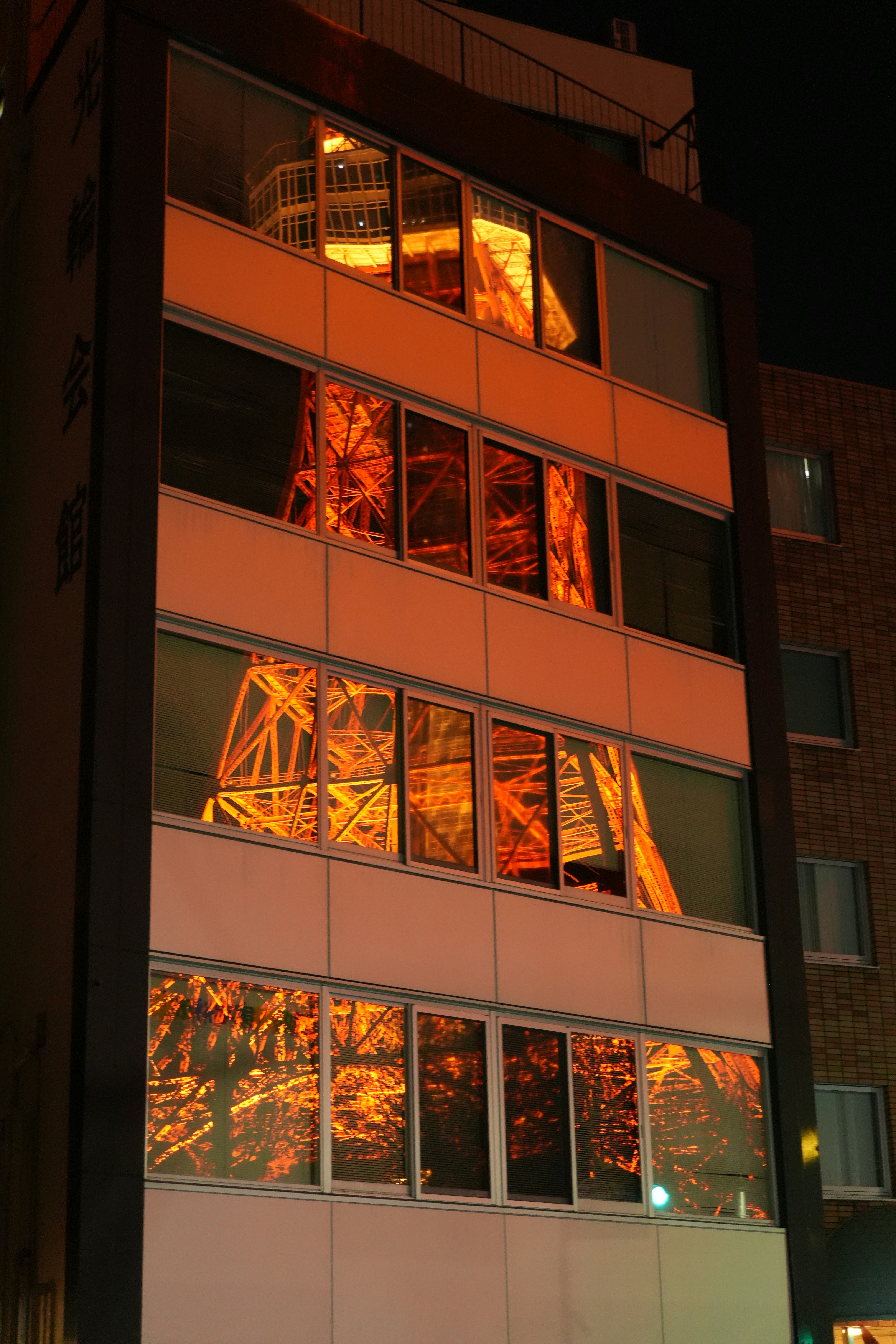 Tokyo Tower illuminated at night, reflected in the windows of a nearby building, showcasing a vibrant interplay of light and structure.