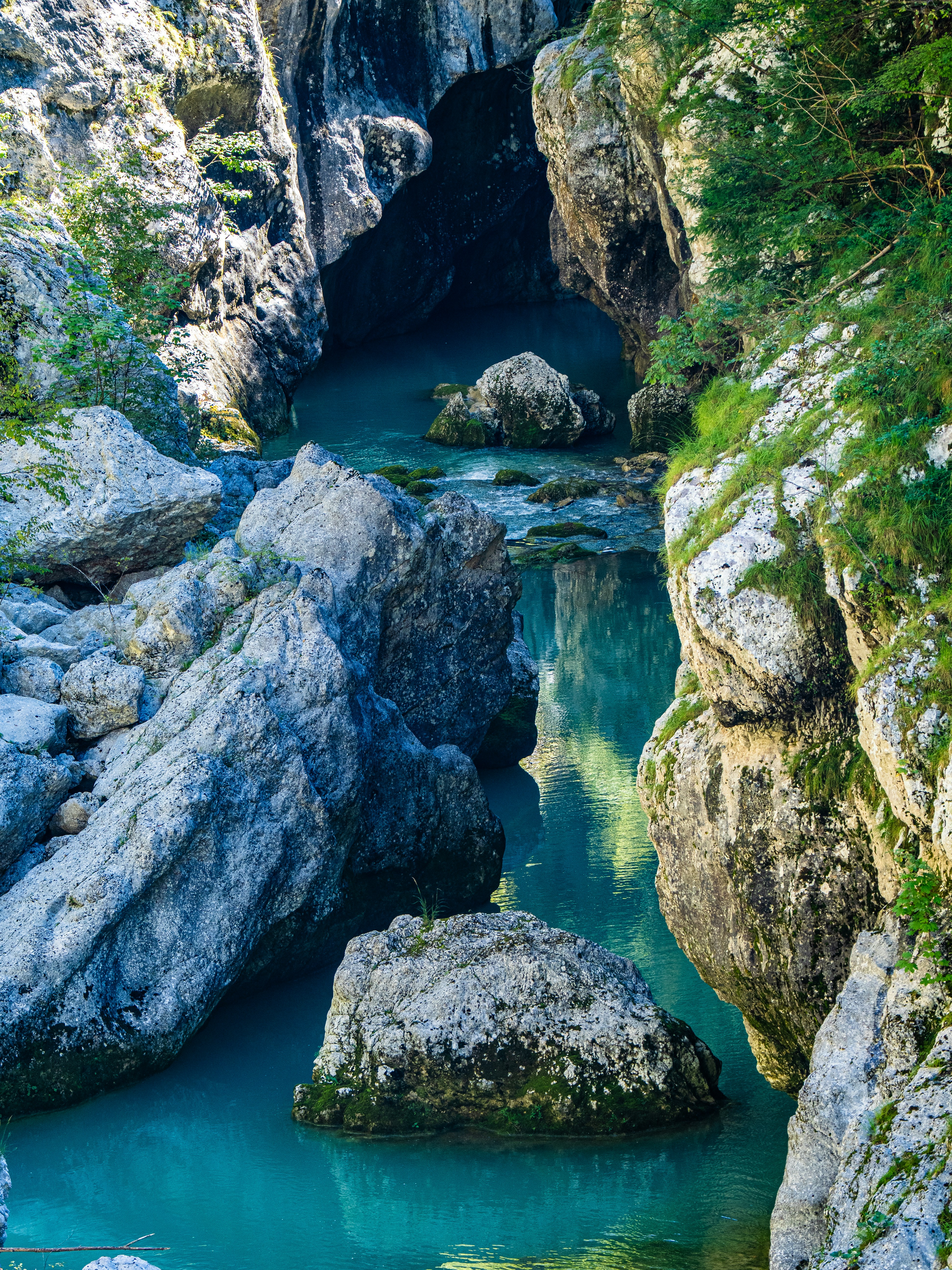 Crystal-clear turquoise waters flowing through rugged rocks and lush greenery, leading into a dark cave entrance.