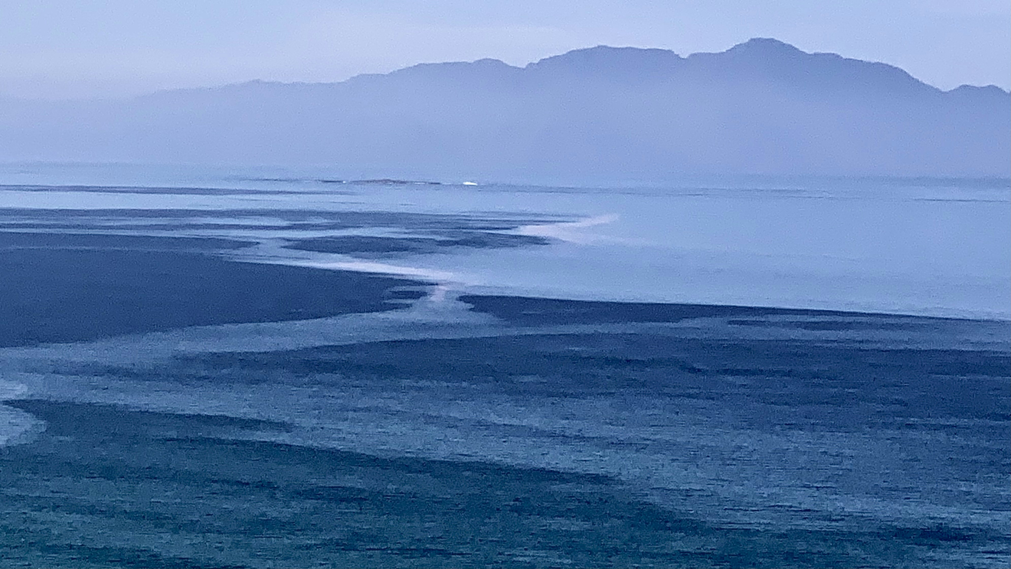 Gentle waves create patterns across the tranquil sea, with distant mountains shrouded in mist. The scene evokes a sense of calm and serenity.