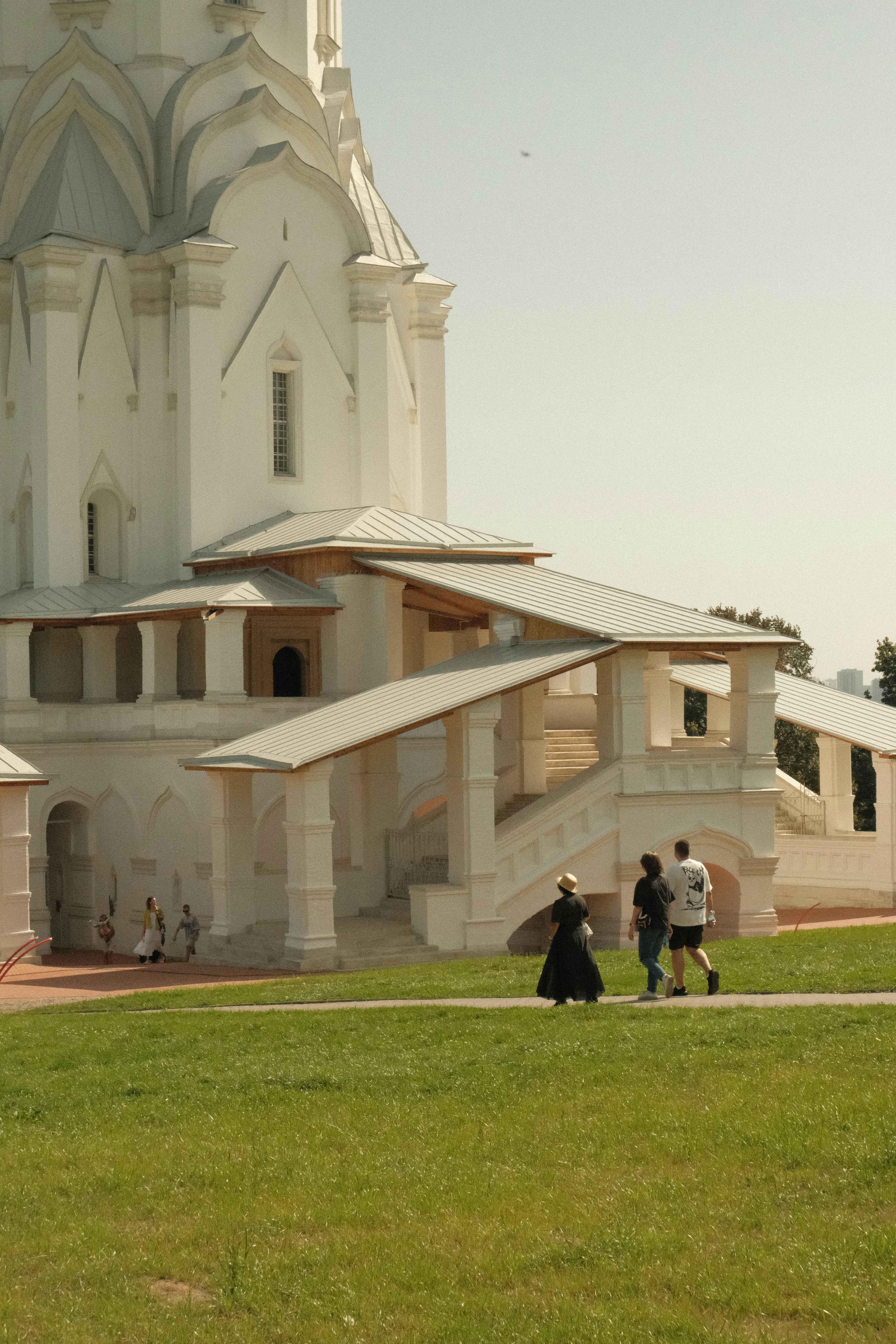 Visitors walking past a grand, white architectural structure with intricate details and multiple levels. The scene captures a blend of nature and history.