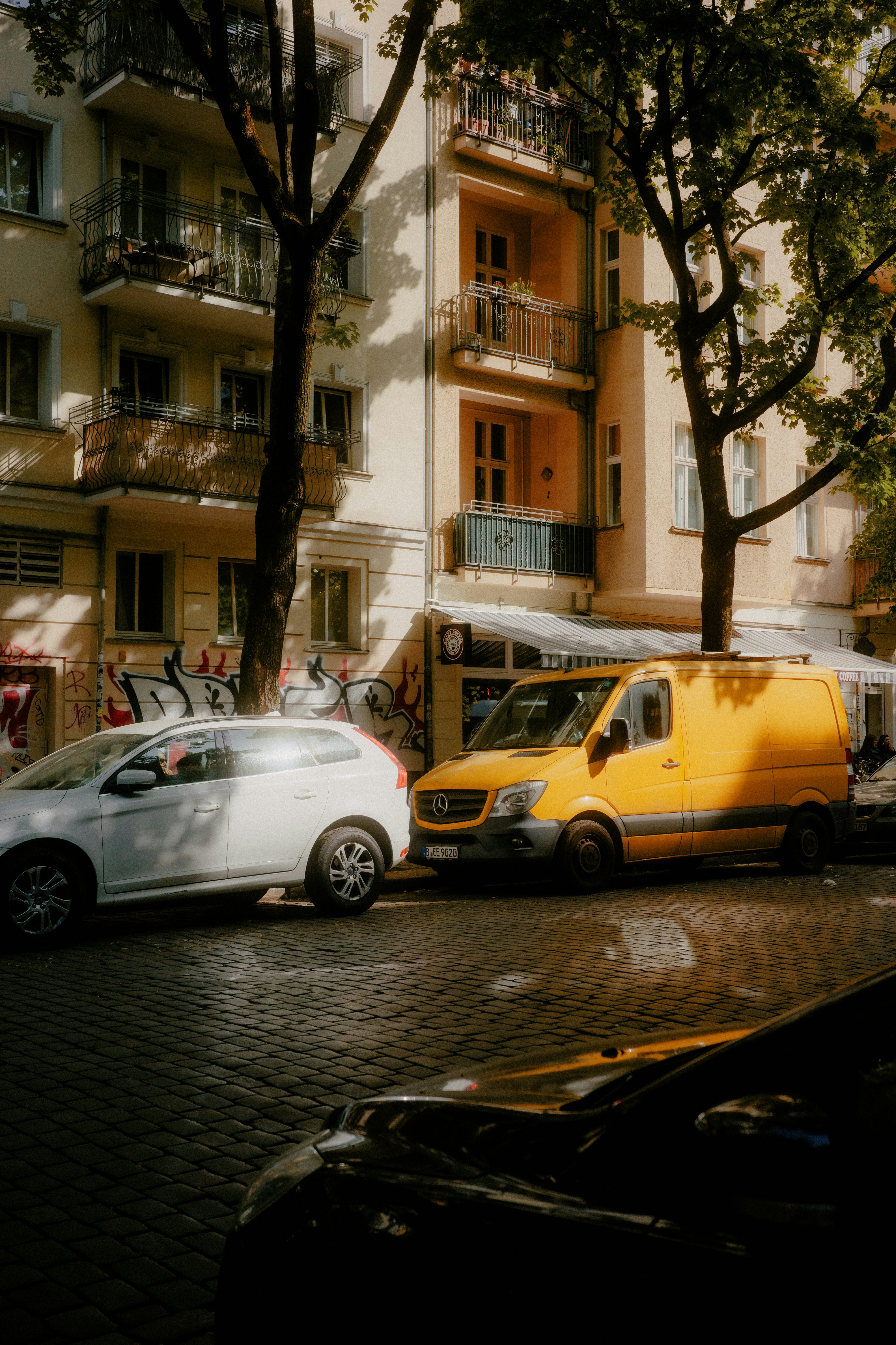 A white SUV and a bright yellow van parked under the dappled sunlight of a tree-lined street, showcasing urban life against a backdrop of graffiti and balconies.