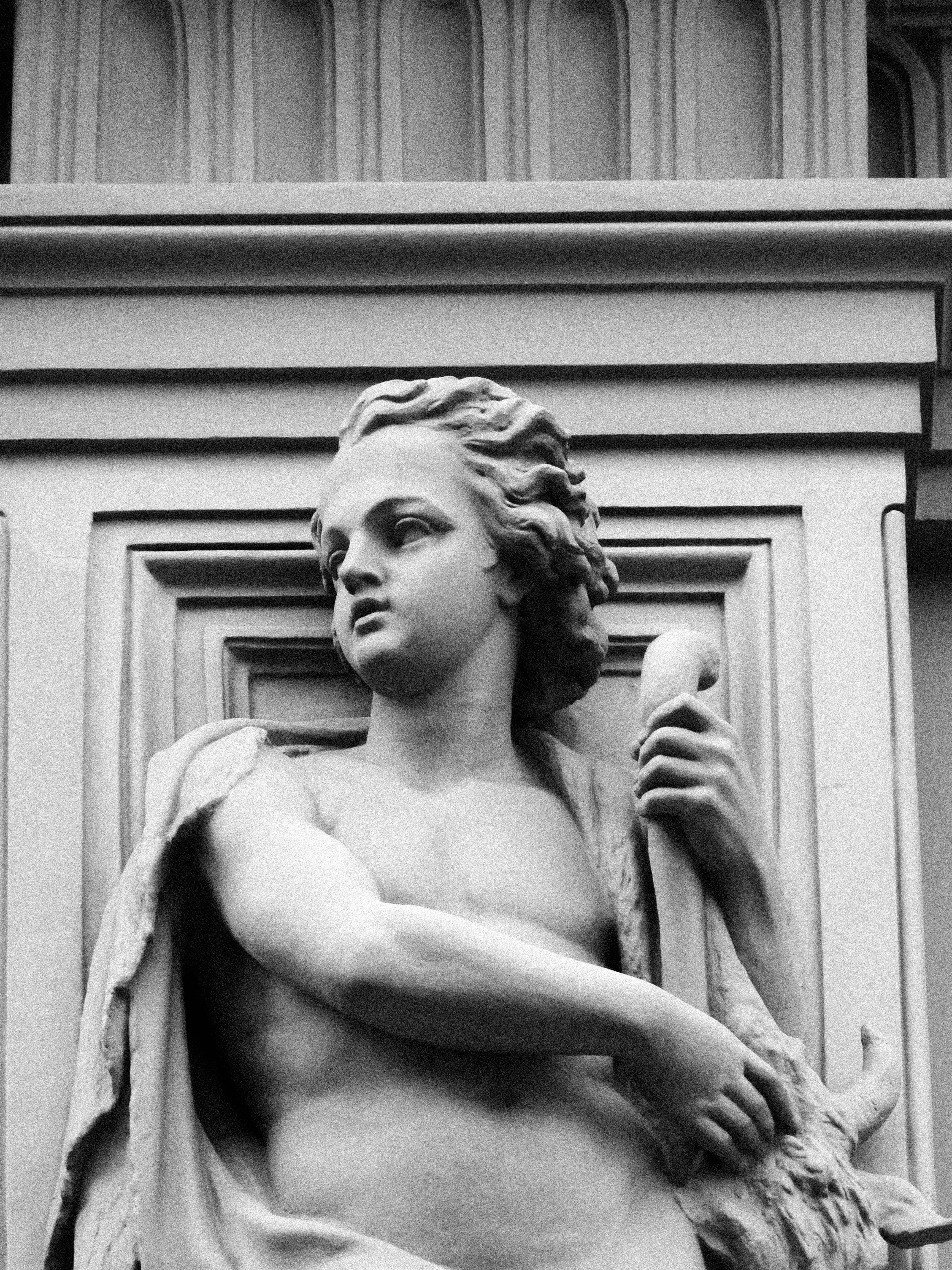 Classical marble sculpture of a figure holding a staff, exuding a sense of strength and serenity against an architectural backdrop.
