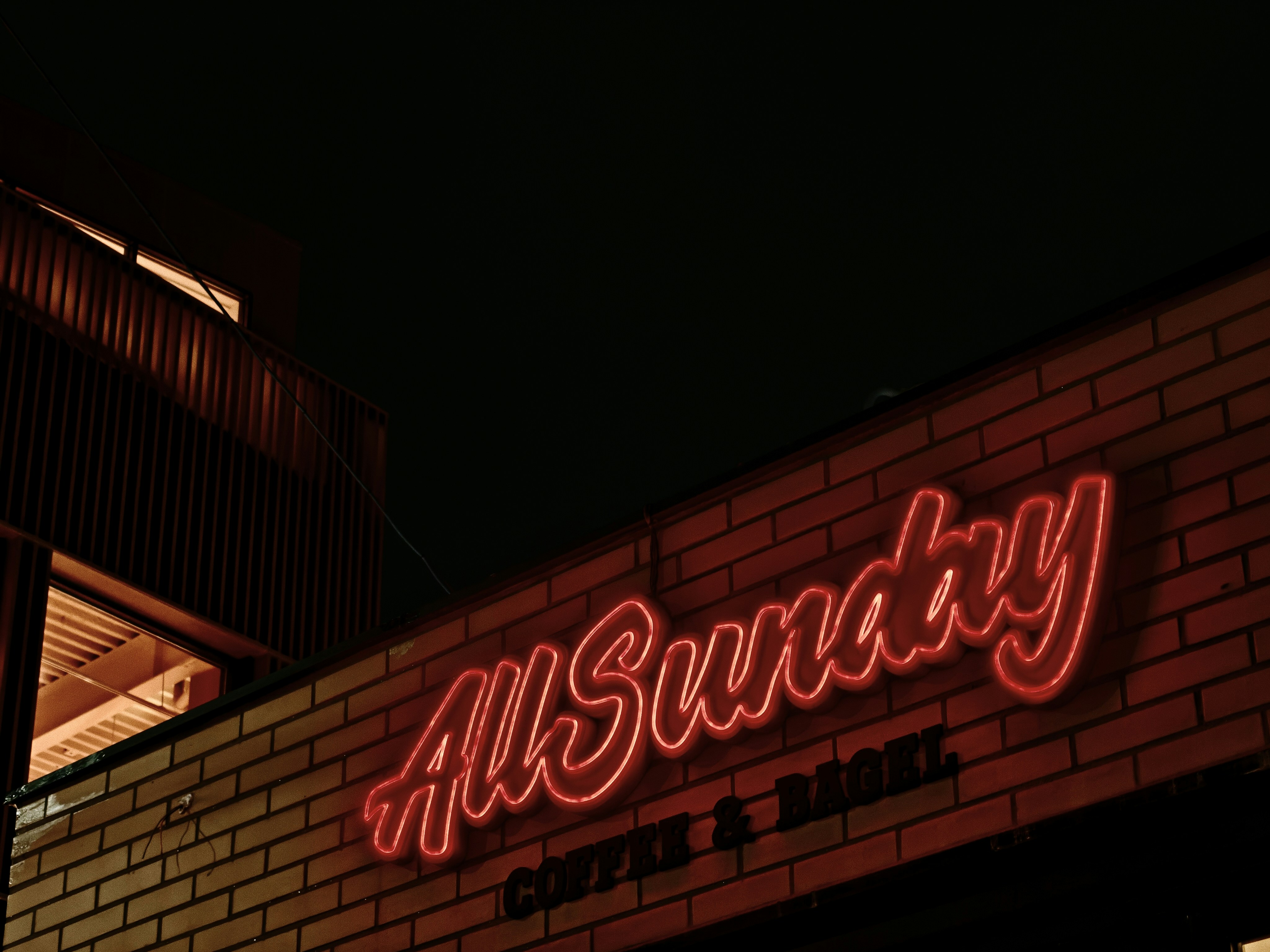 Illuminated neon sign for AllSunday Coffee & Bagel on a brick wall at night, highlighting the vibrant atmosphere of the café.