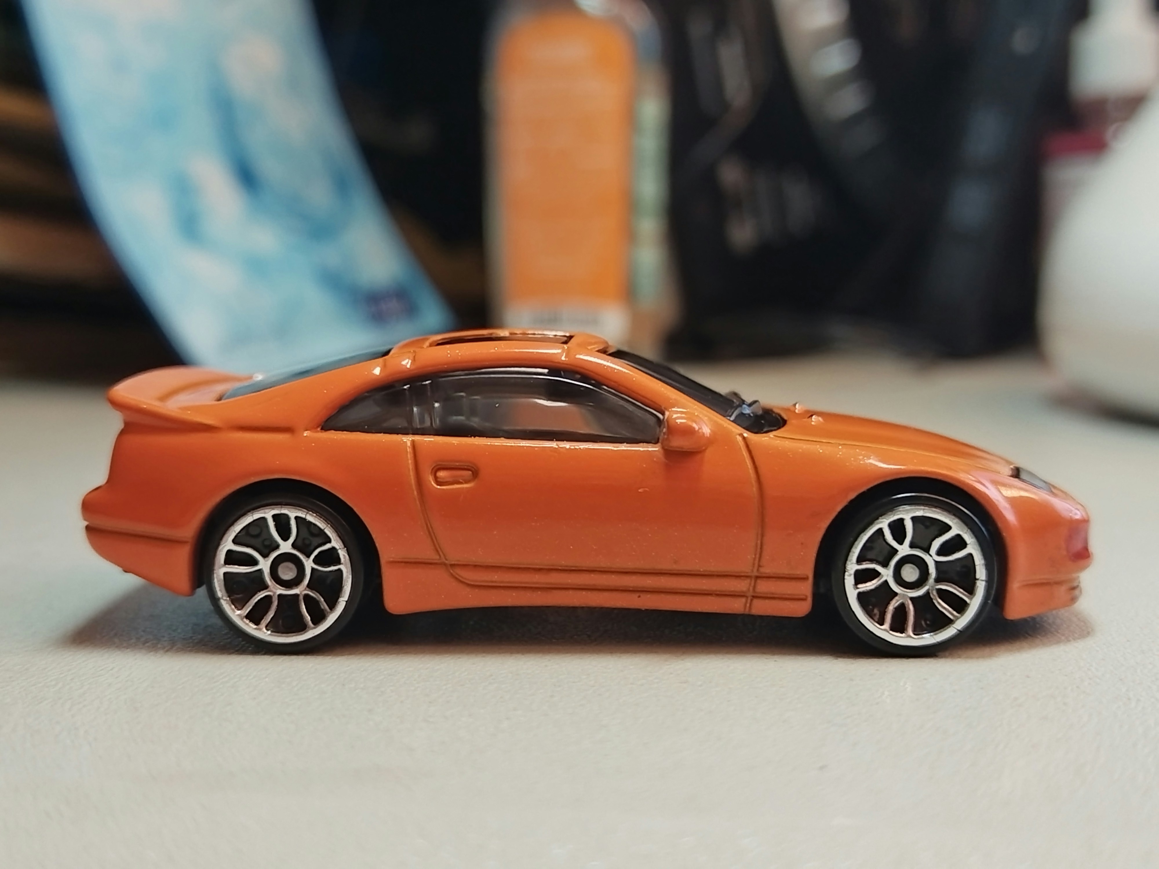 Detailed orange toy car model with sleek design and sporty wheels, set against a blurred background of everyday objects.