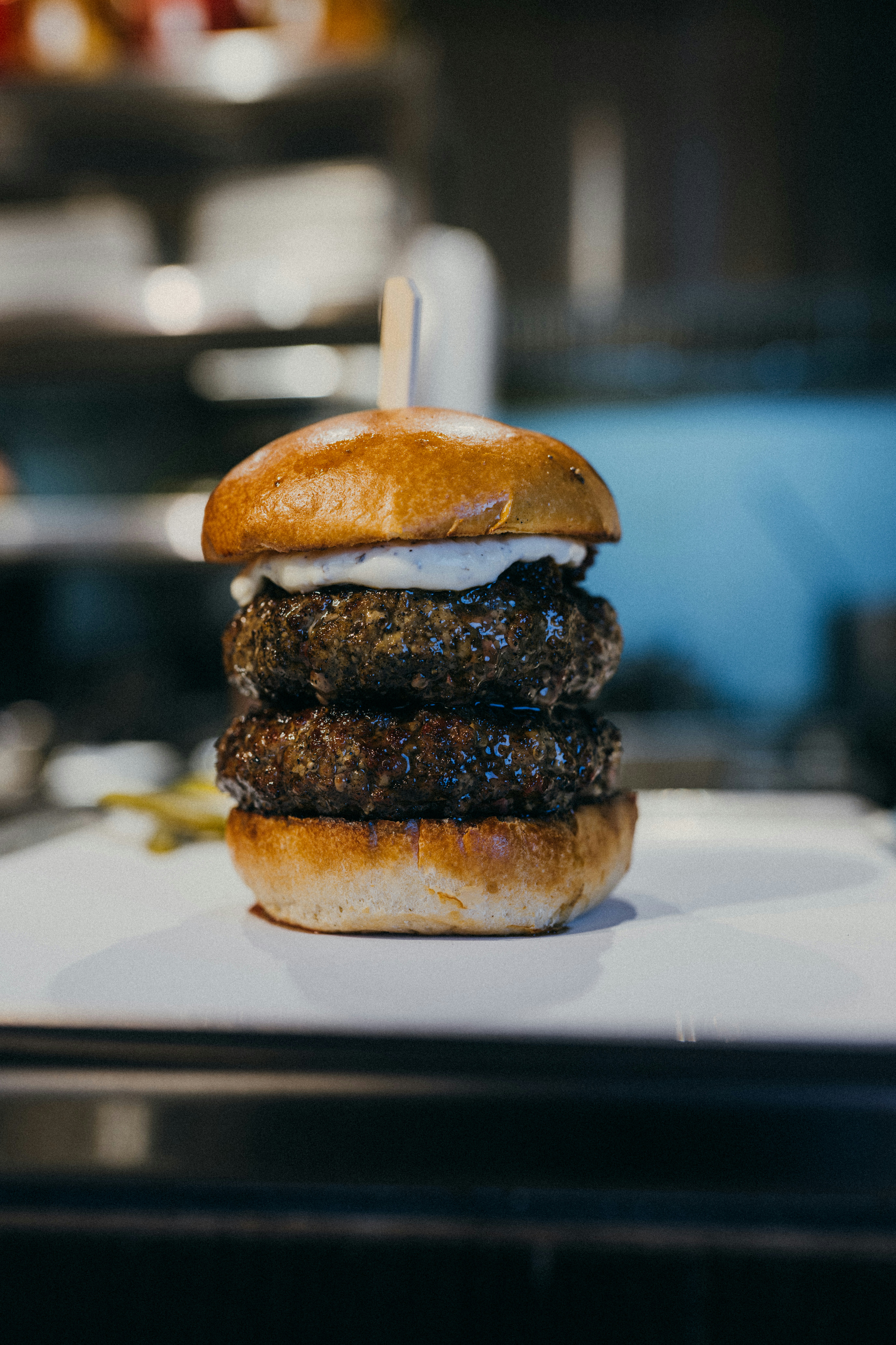 A towering burger featuring three juicy patties, topped with a golden bun and creamy sauce, set against a blurred kitchen backdrop.