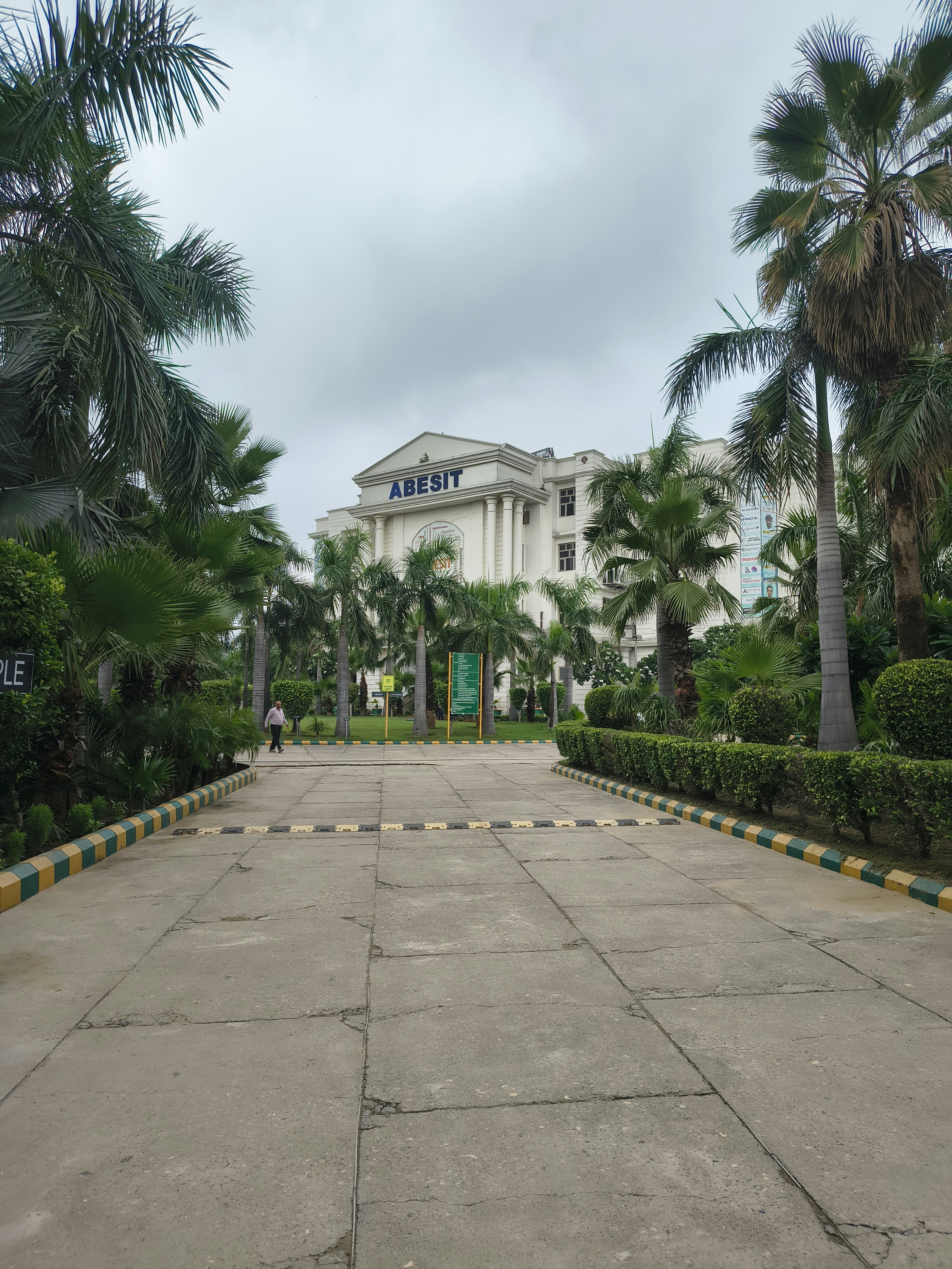 A grand, classical-style building with "ABESIT" written on its facade, surrounded by lush green palm trees and manicured gardens. The image captures the main entrance, with a paved road leading up to the building under a cloudy sky, suggesting a professional and academic environment. | Modern building entrance with palm trees and walkway