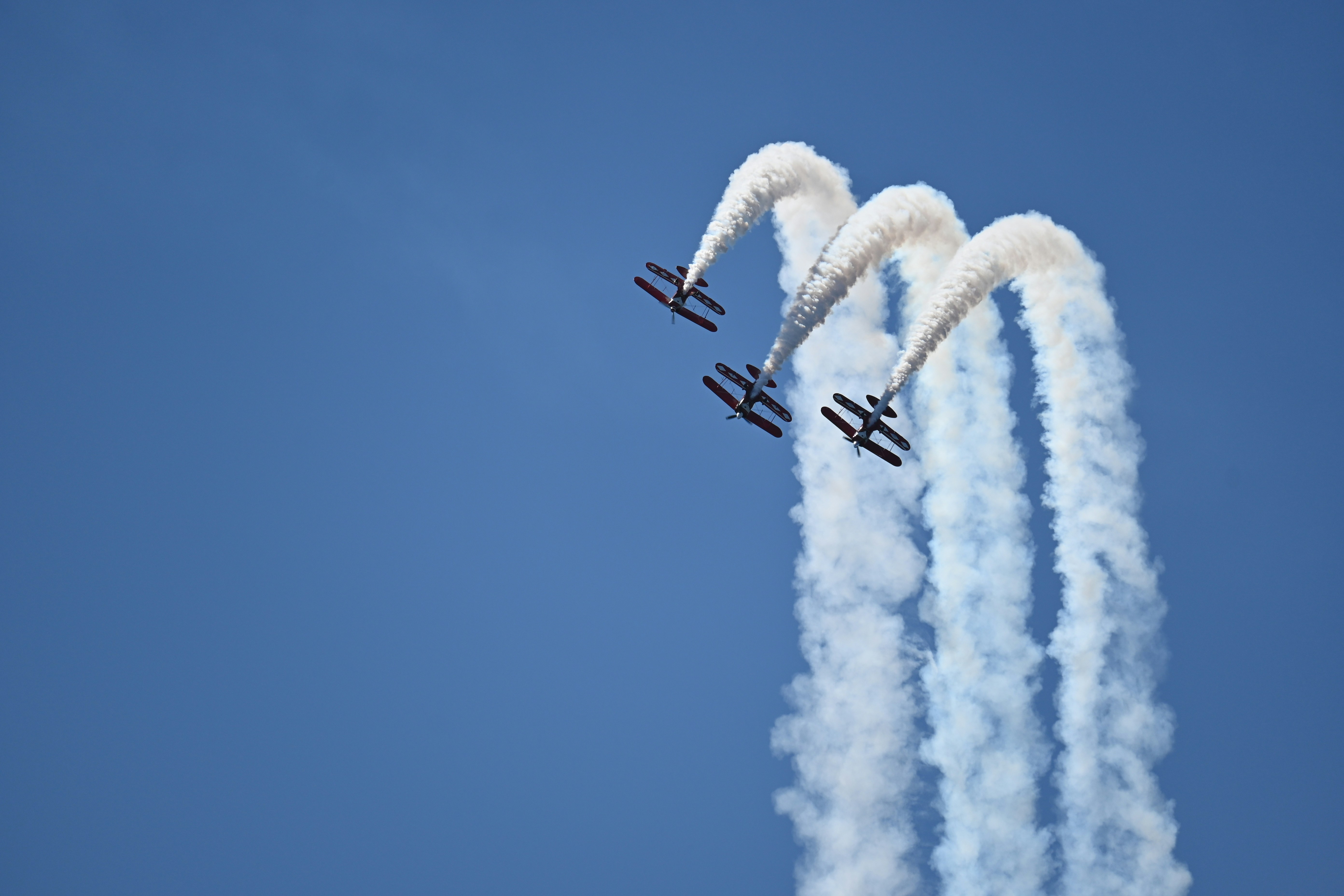 Three red biplanes are flying in a close formation, performing an acrobatic maneuver against a clear blue sky. Each plane leaves a thick, white smoke trail, which forms three parallel inverted loops. | Three planes perform aerobatics, leaving white smoke trails.