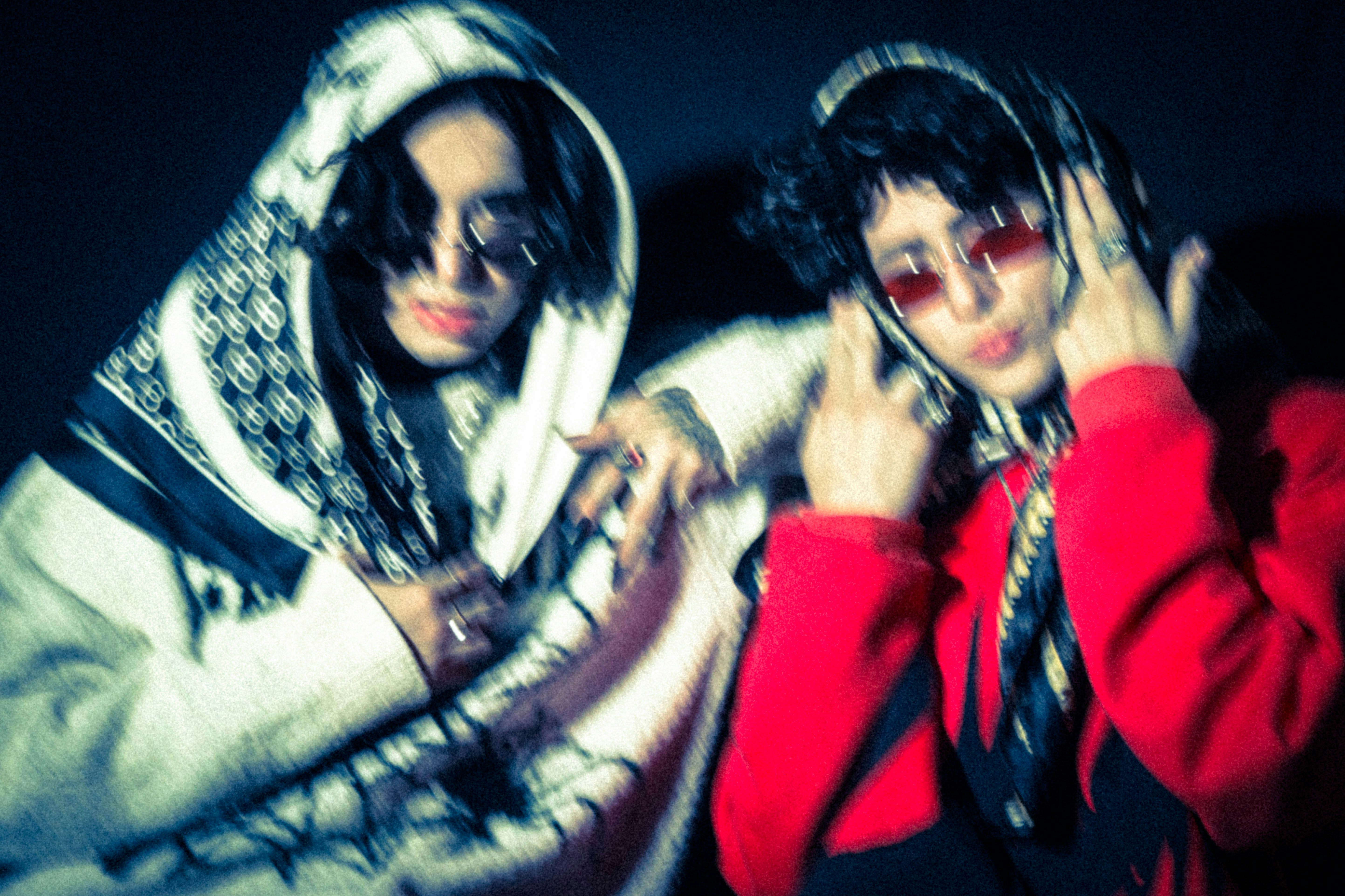 Two individuals in stylish attire, shrouded in scarves and sunglasses, pose against a dark backdrop, exuding an air of intrigue.