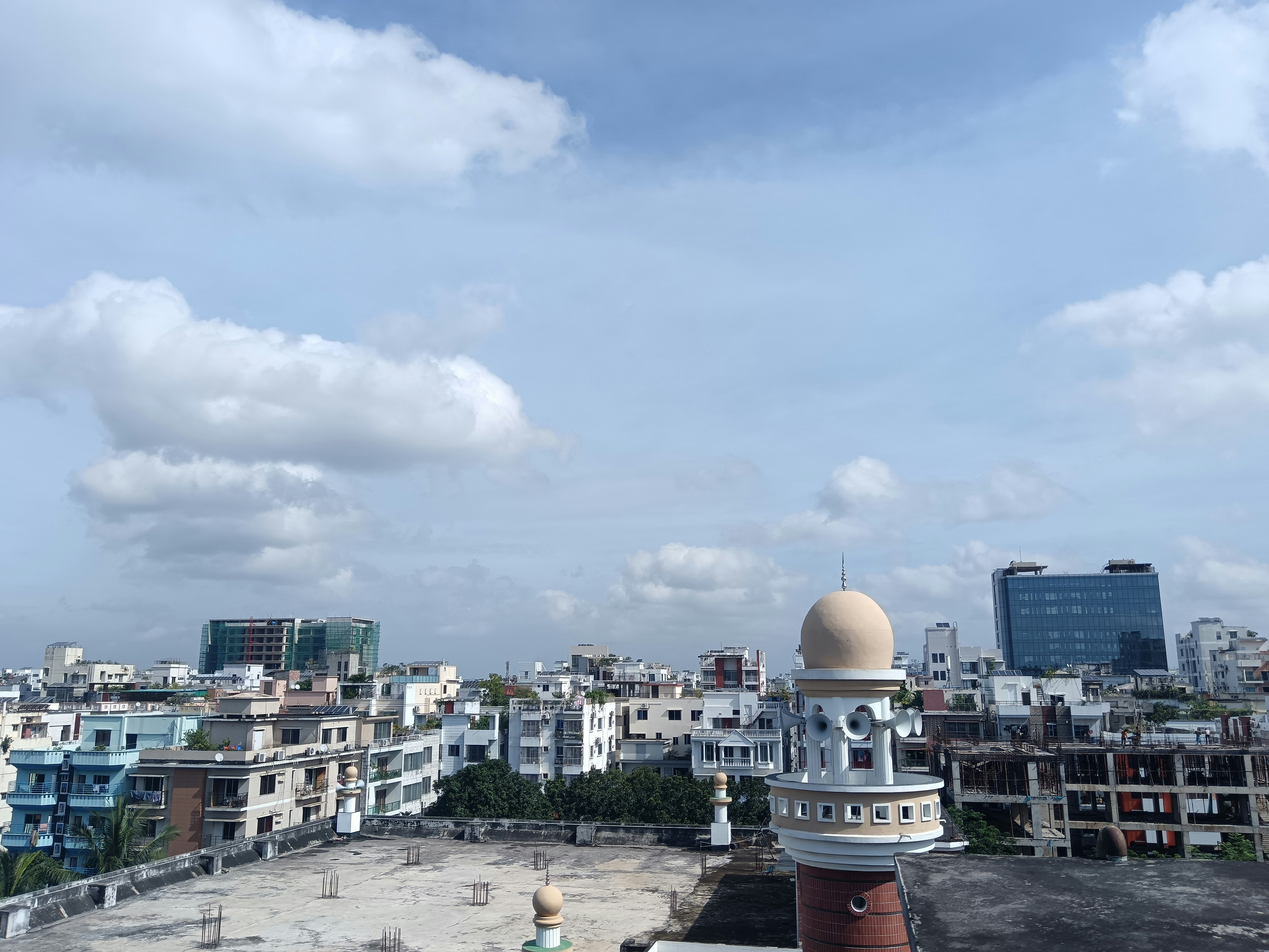 City skyline featuring a mix of modern and traditional architecture, with a prominent dome structure in the foreground. Fluffy clouds drift across a bright blue sky.