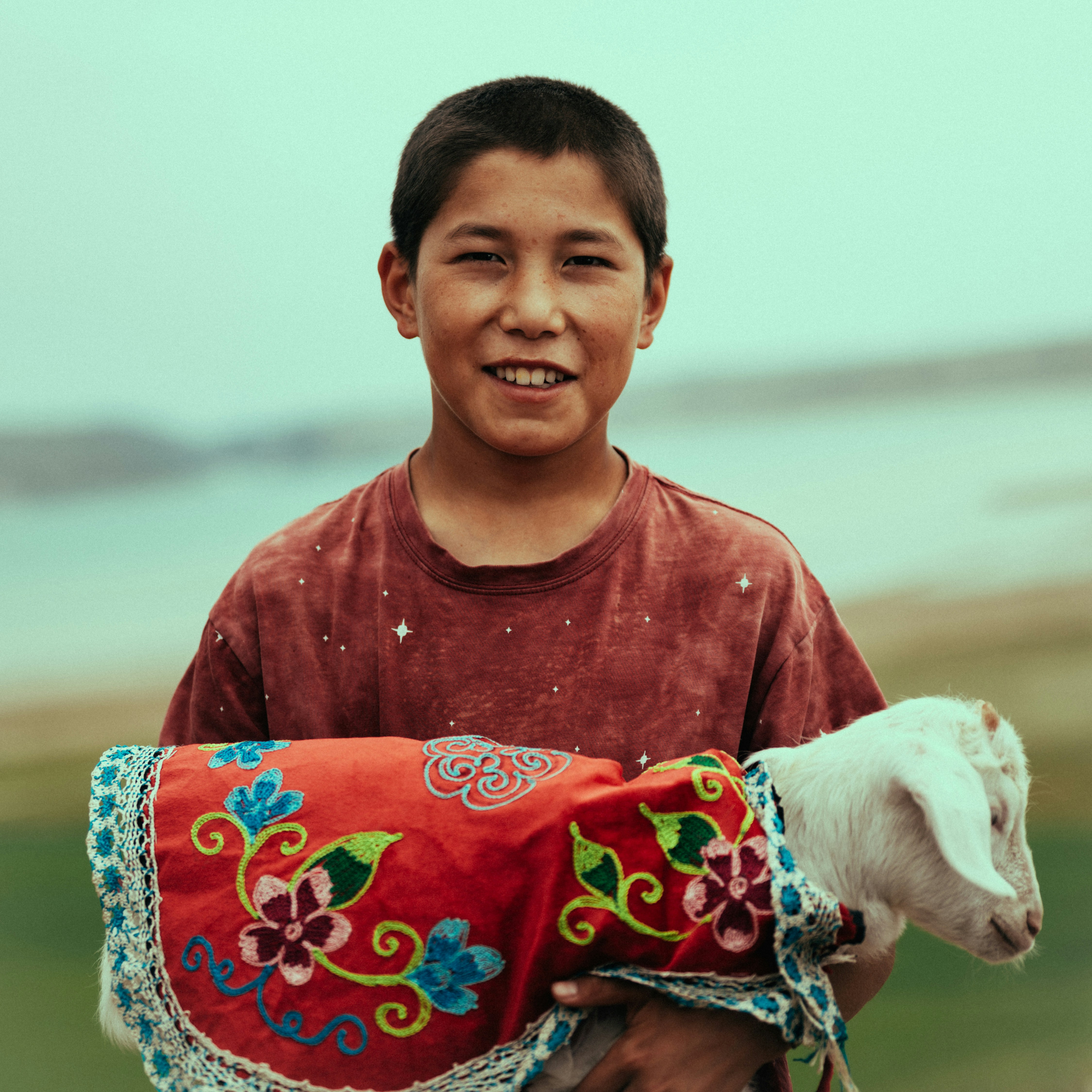 Young boy wearing a colorful garment holds a goat wrapped in a decorative blanket against a serene backdrop. 