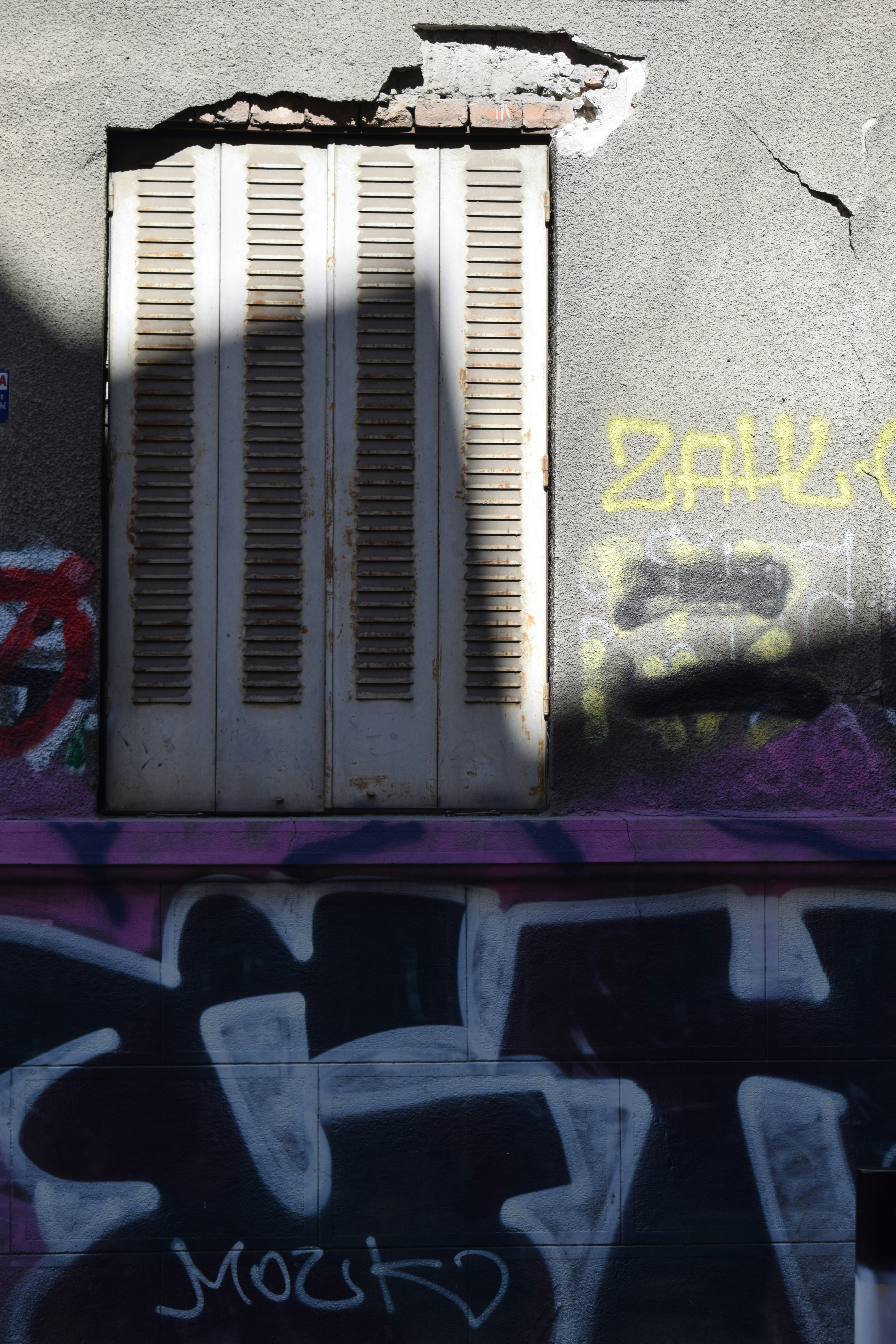 Weathered shuttered window casting shadows on a graffiti-covered wall, revealing urban decay and artistic expression.