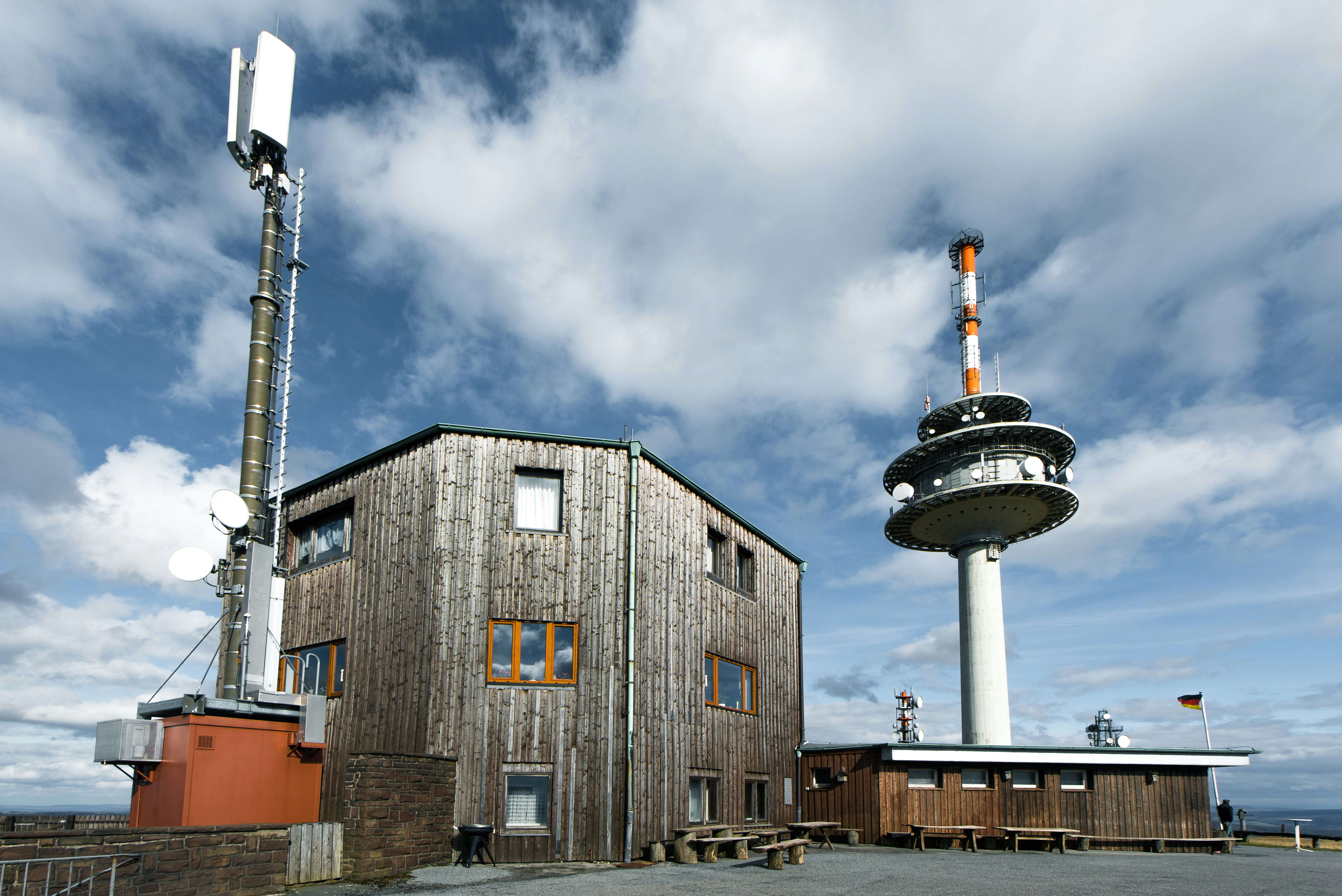 A wooden observation building stands beside a modern communication tower against a backdrop of dynamic clouds. The structure's rustic charm contrasts with the sleek technology above.