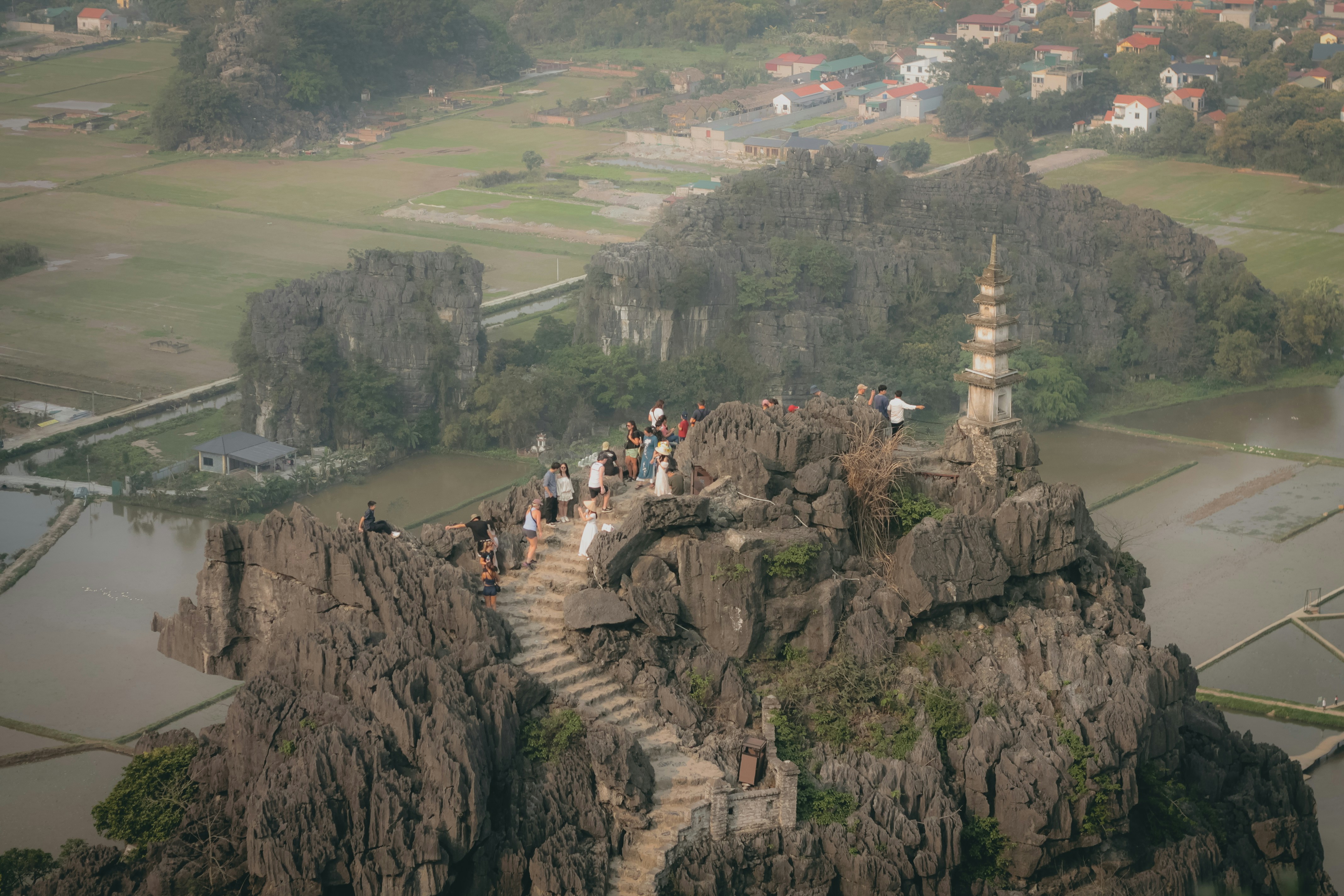 Visitors gather at the peak of a rocky outcrop, surrounded by lush fields and a distant pagoda. A serene landscape unfolds below.