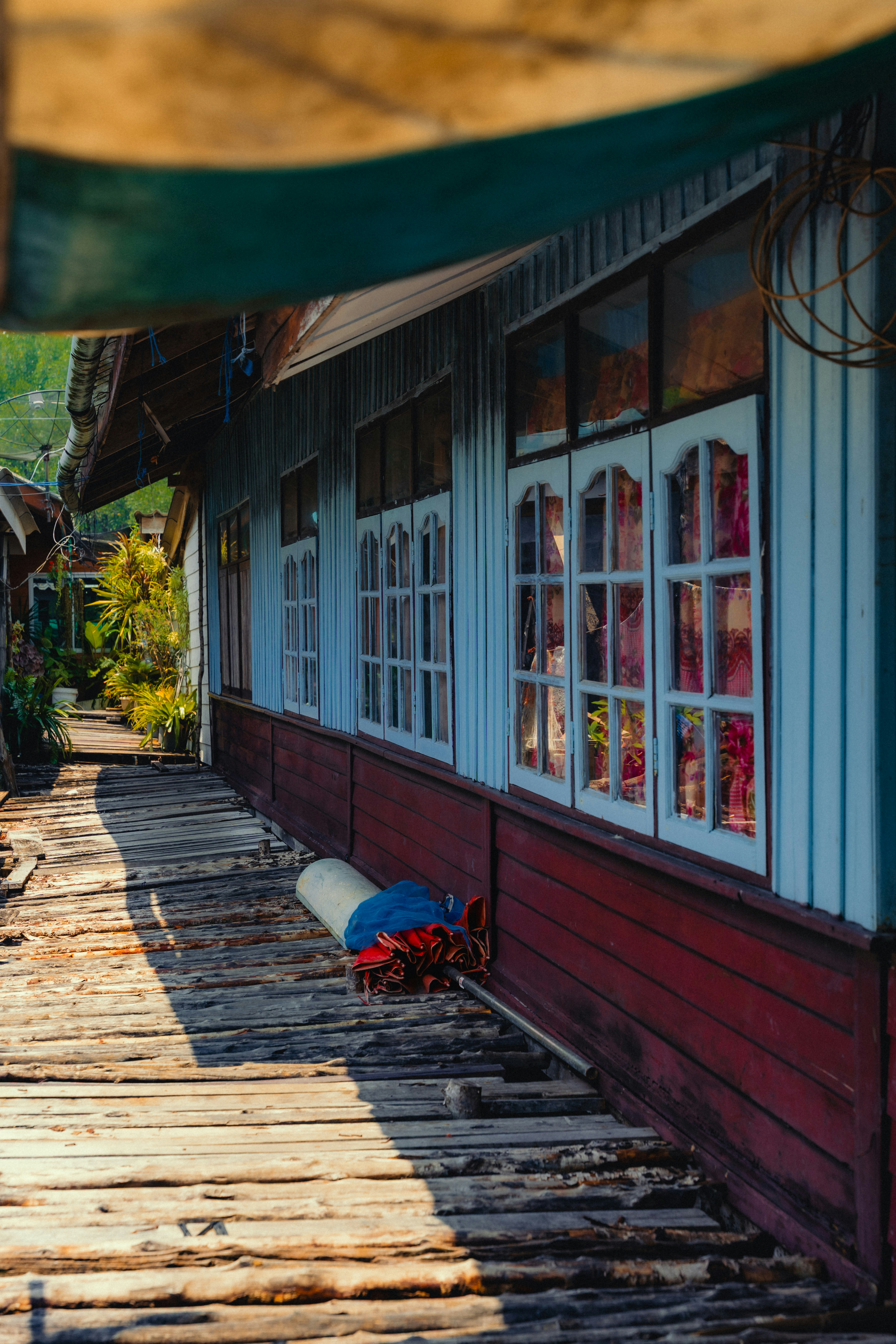 A traditional wooden walkway leading past colorful houses with windows reflecting sunlight. | Wooden walkway beside a light blue building