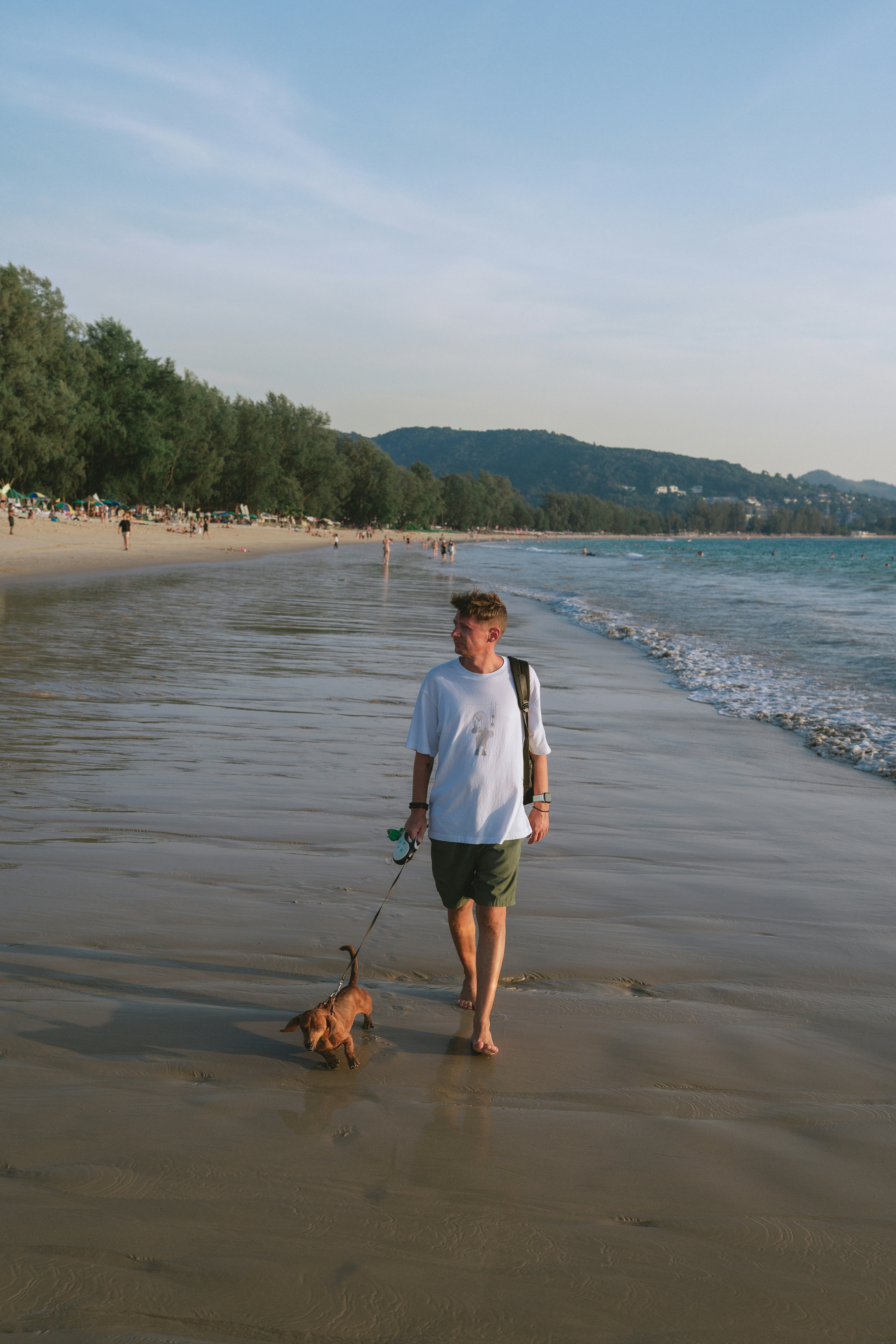 A man walking barefoot on the beach with a small dog on a leash, evening light. | Man walking dog on a sandy beach