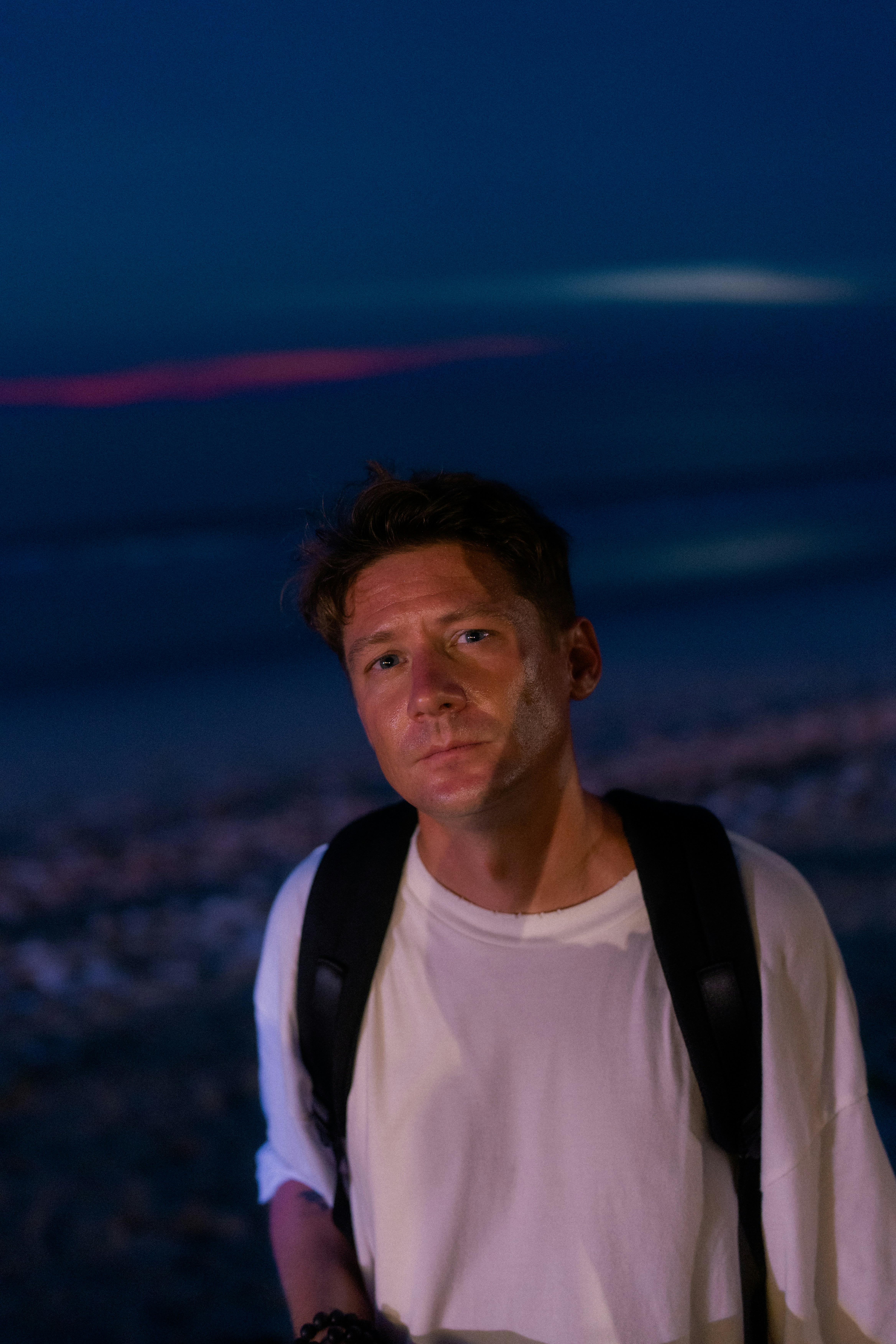Portrait of a man at night on the beach, wearing a white shirt and backpack, moody lighting. | Man with backpack looking at camera at camera at night