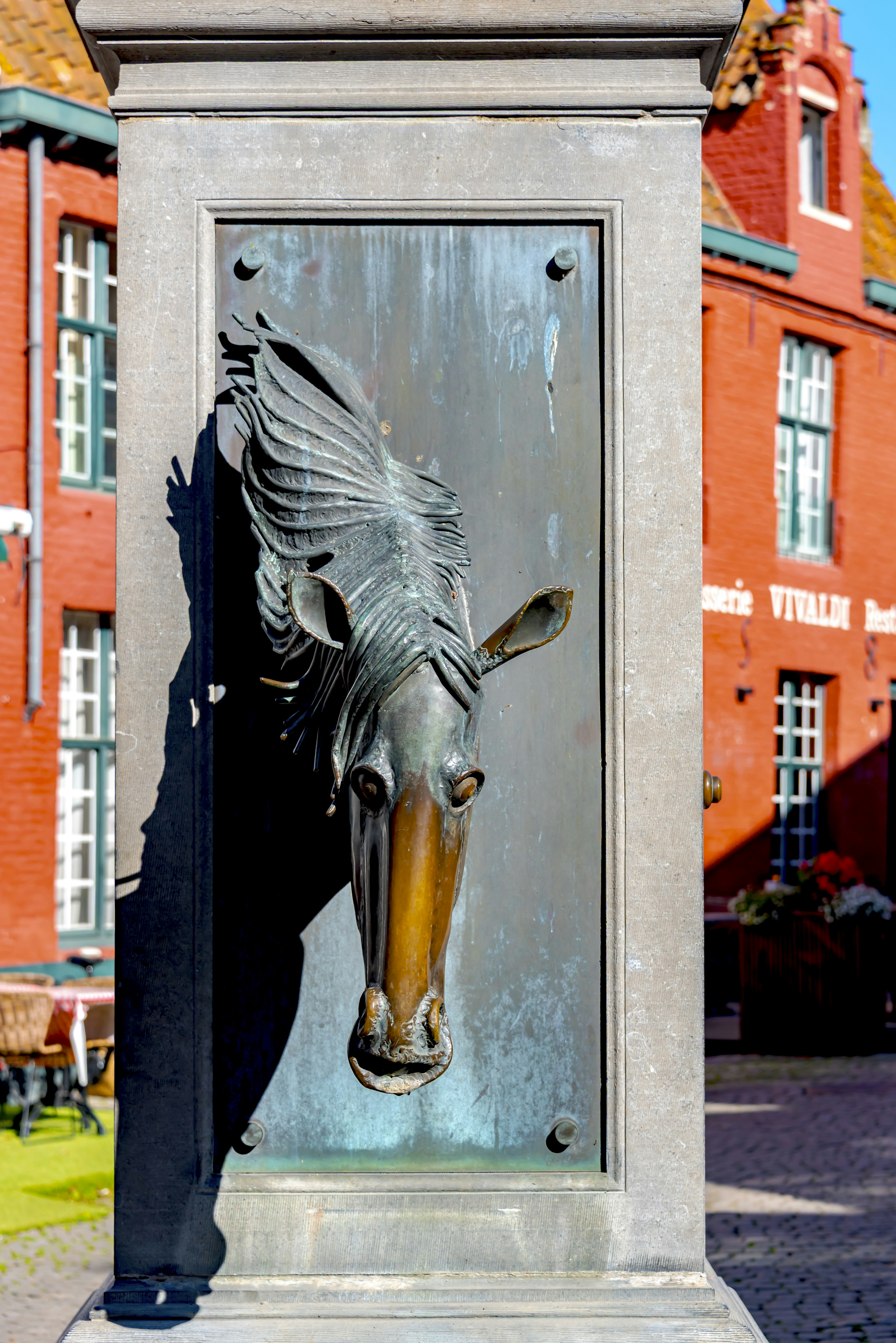 Bronze sculpture of a horse's head mounted on a stone pillar, set against a backdrop of red-brick buildings.