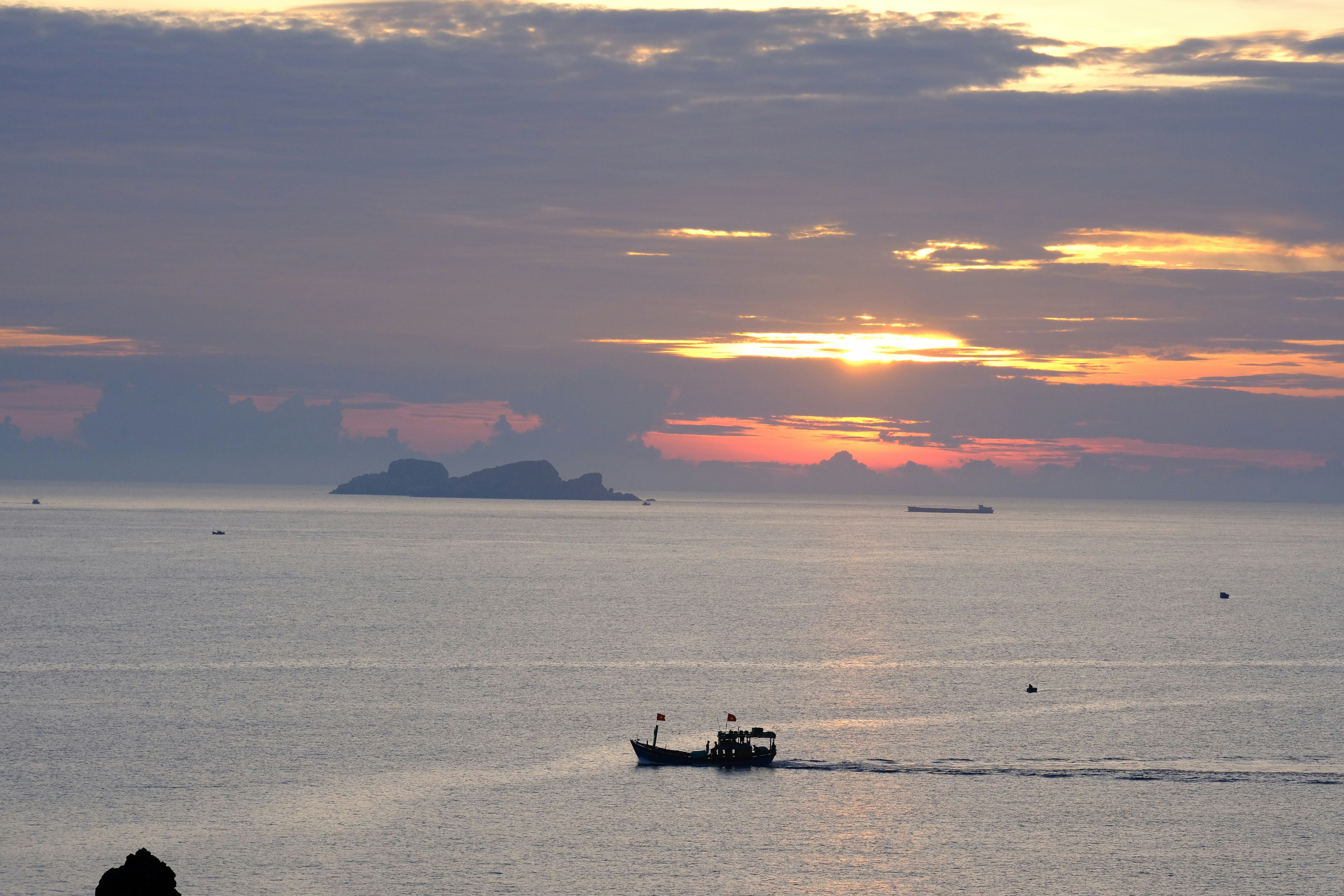 A fishing boat glides across a tranquil sea at sunset, with distant islands silhouetted against the colorful sky.
