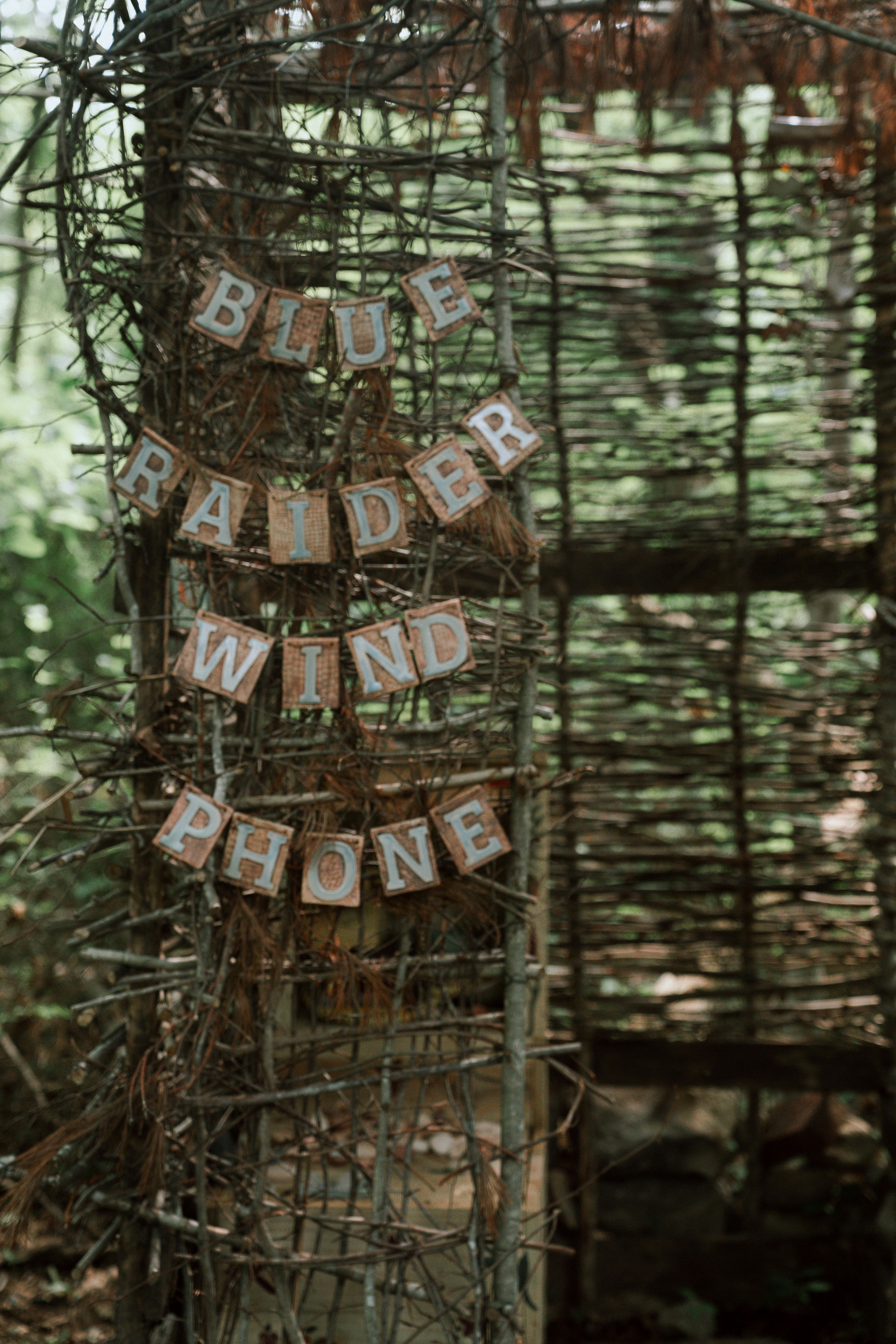 A rustic installation featuring wooden letters spelling 'BLUE RAIDER WIND PHONE' amidst a backdrop of intertwined branches and greenery.