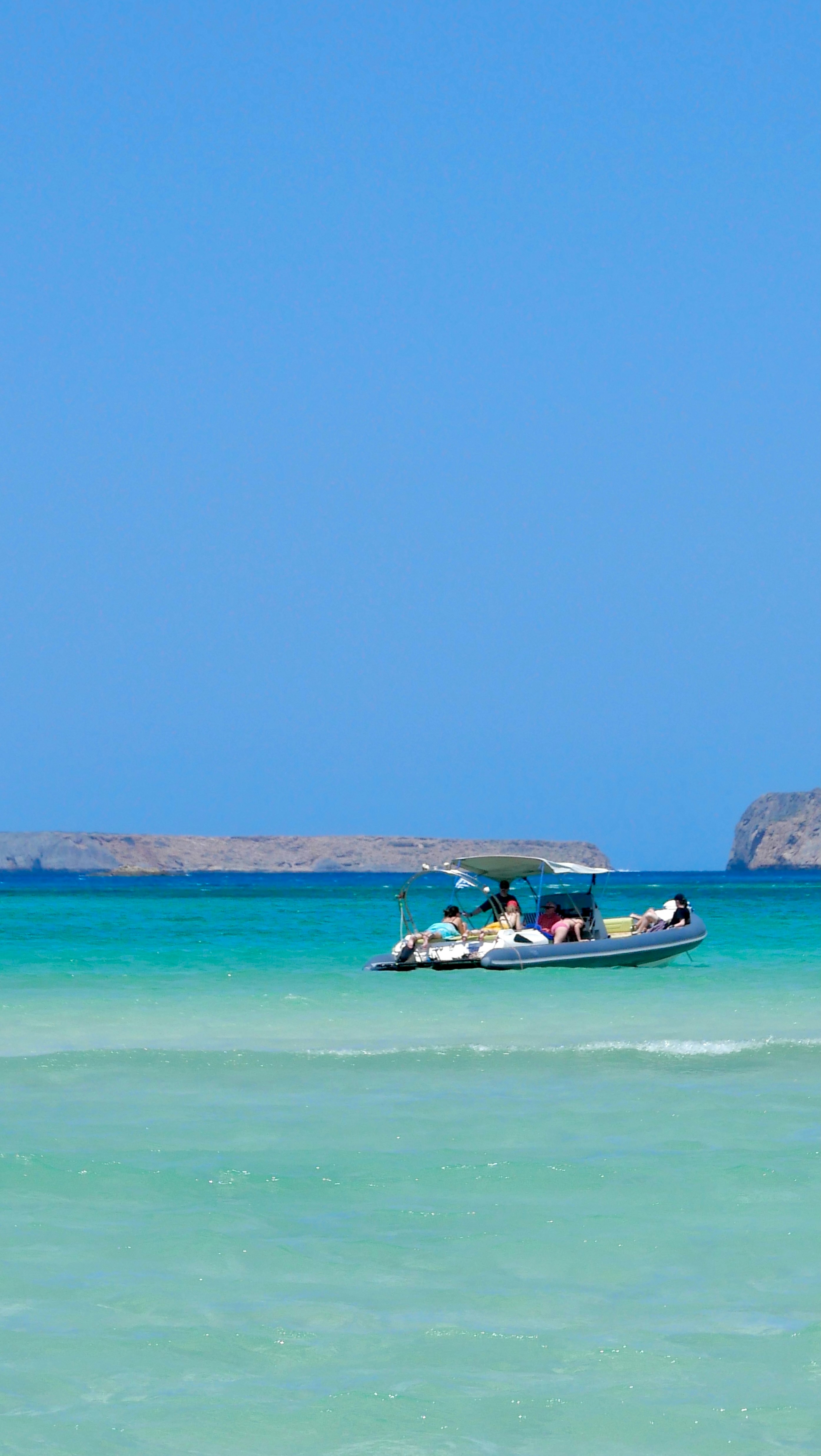 A boat carrying passengers glides through crystal-clear turquoise waters, framed by distant rocky shores under a clear blue sky.