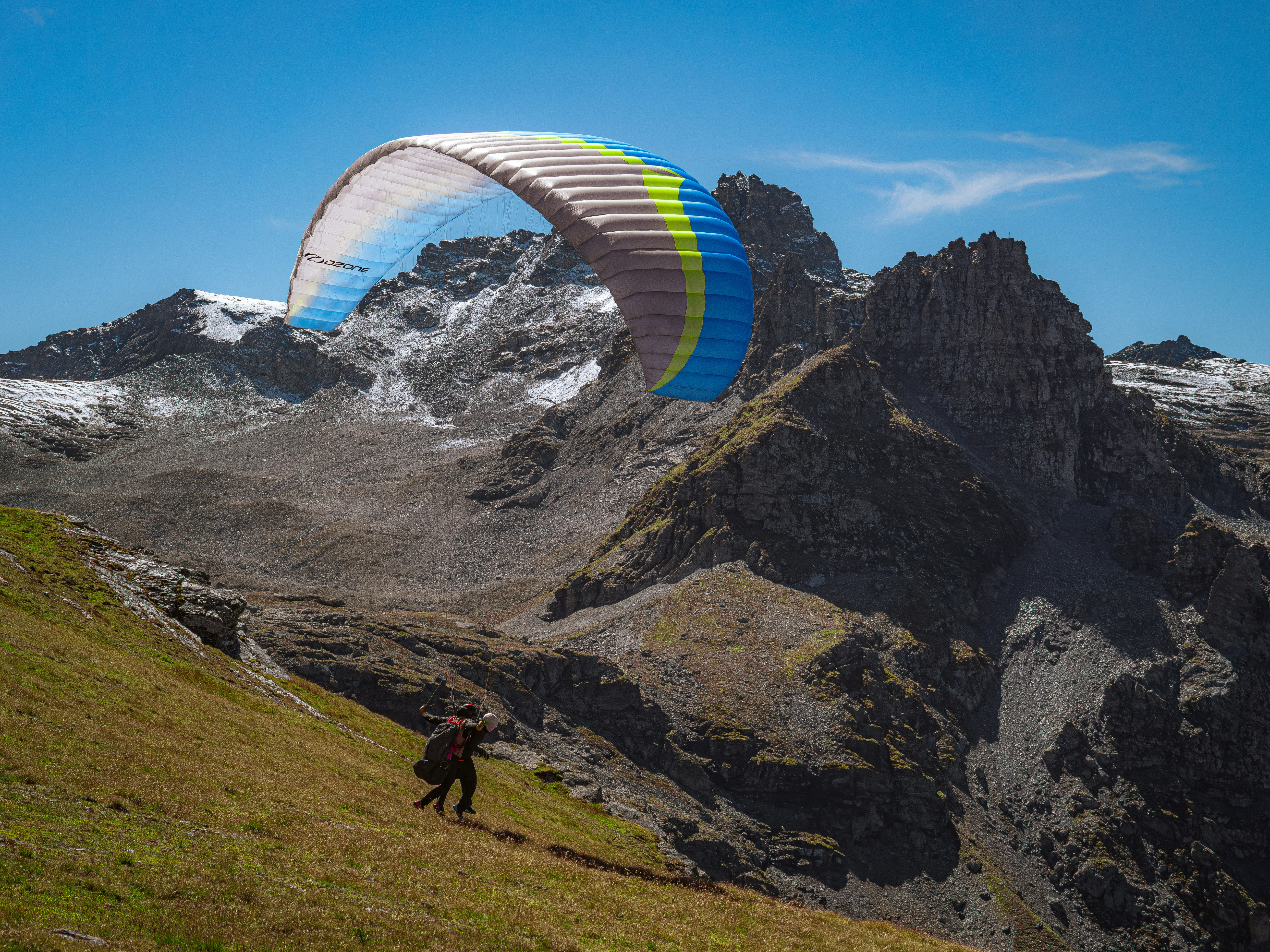 Paraglider launching from a grassy slope, framed by majestic mountains under a clear blue sky.