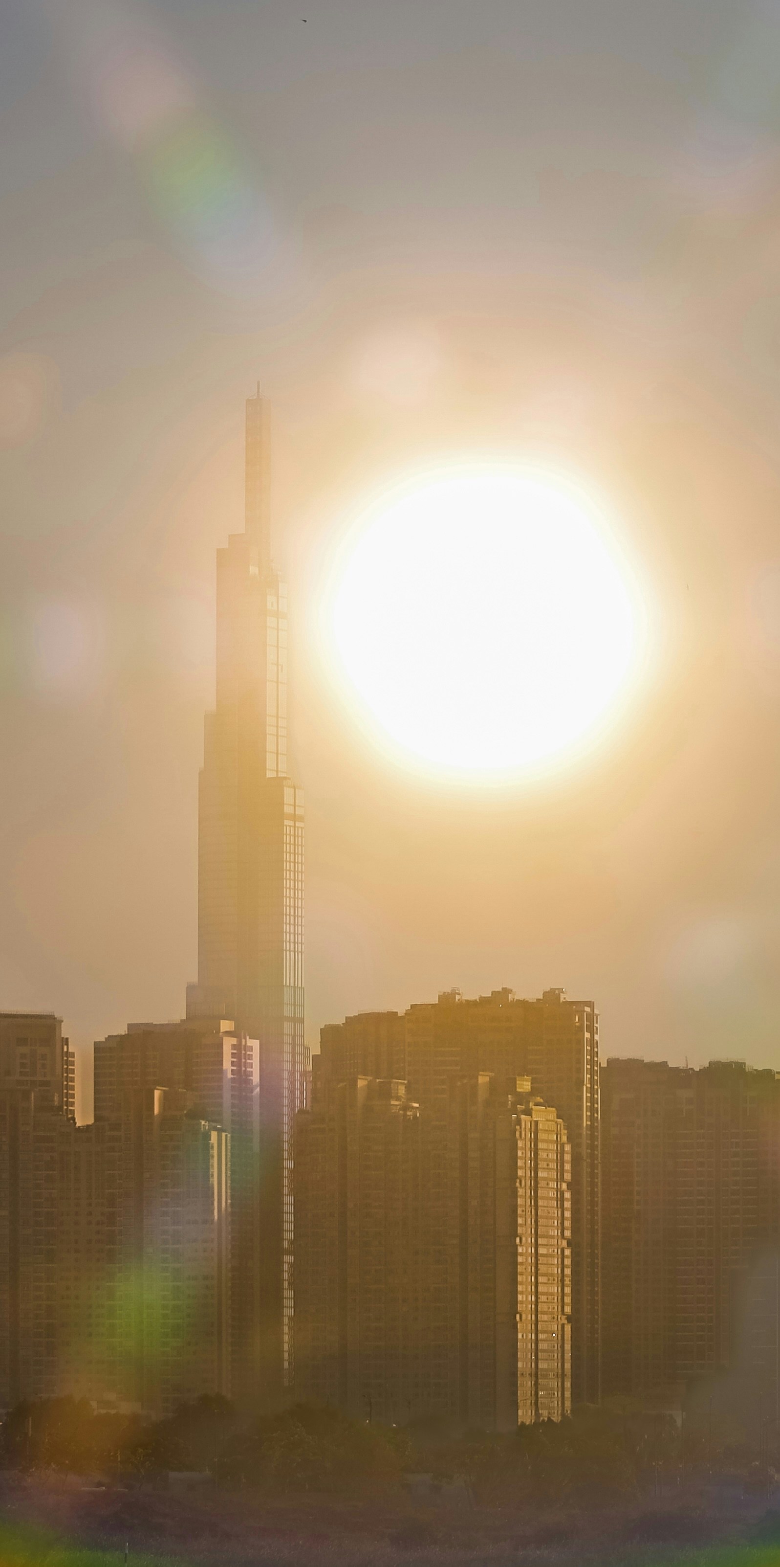Tall skyscraper against a bright sun and city buildings