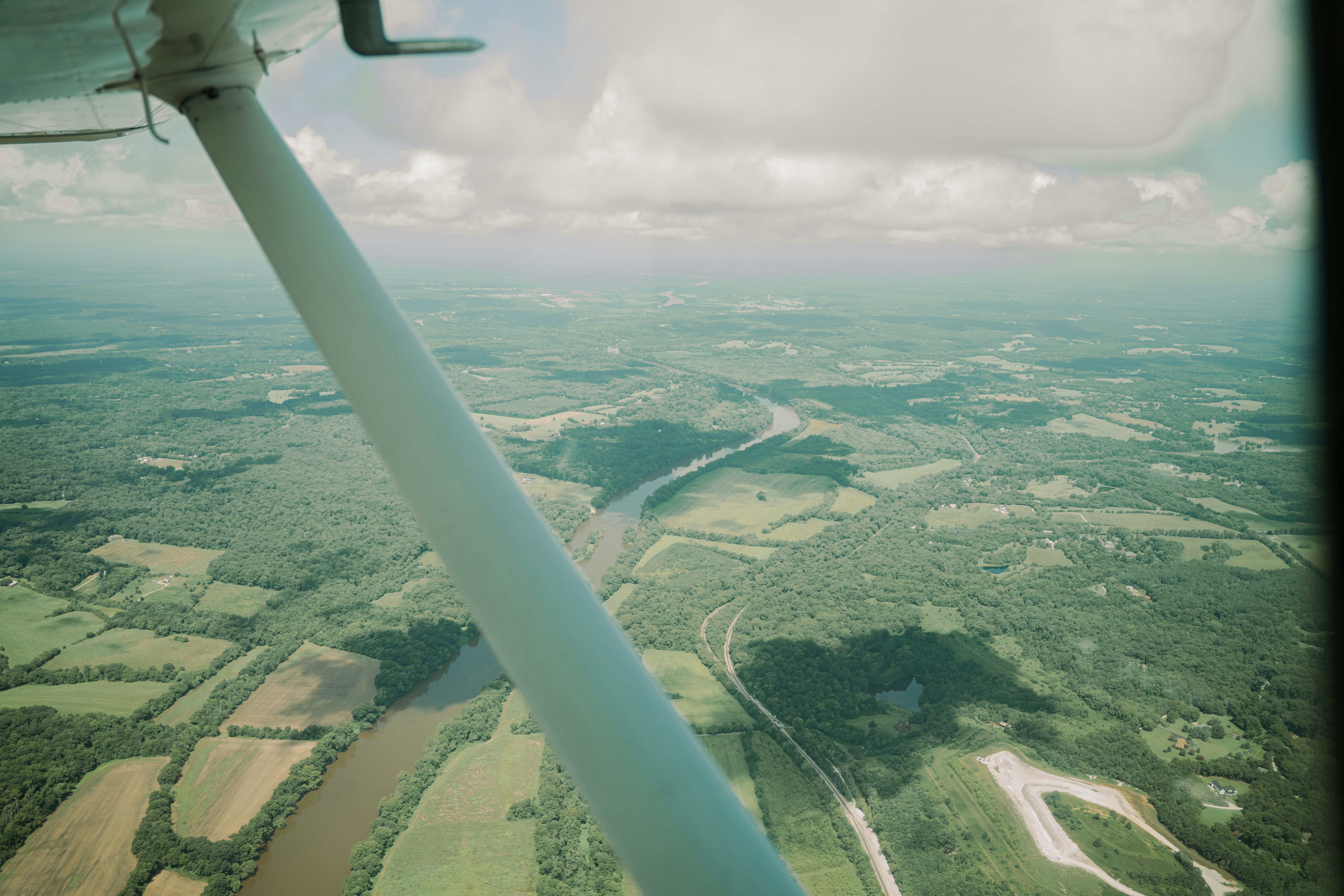Aerial view of lush green landscapes intersected by a winding river, with a wing of an aircraft partially visible. The scene captures the harmony of nature's contours and waterways.