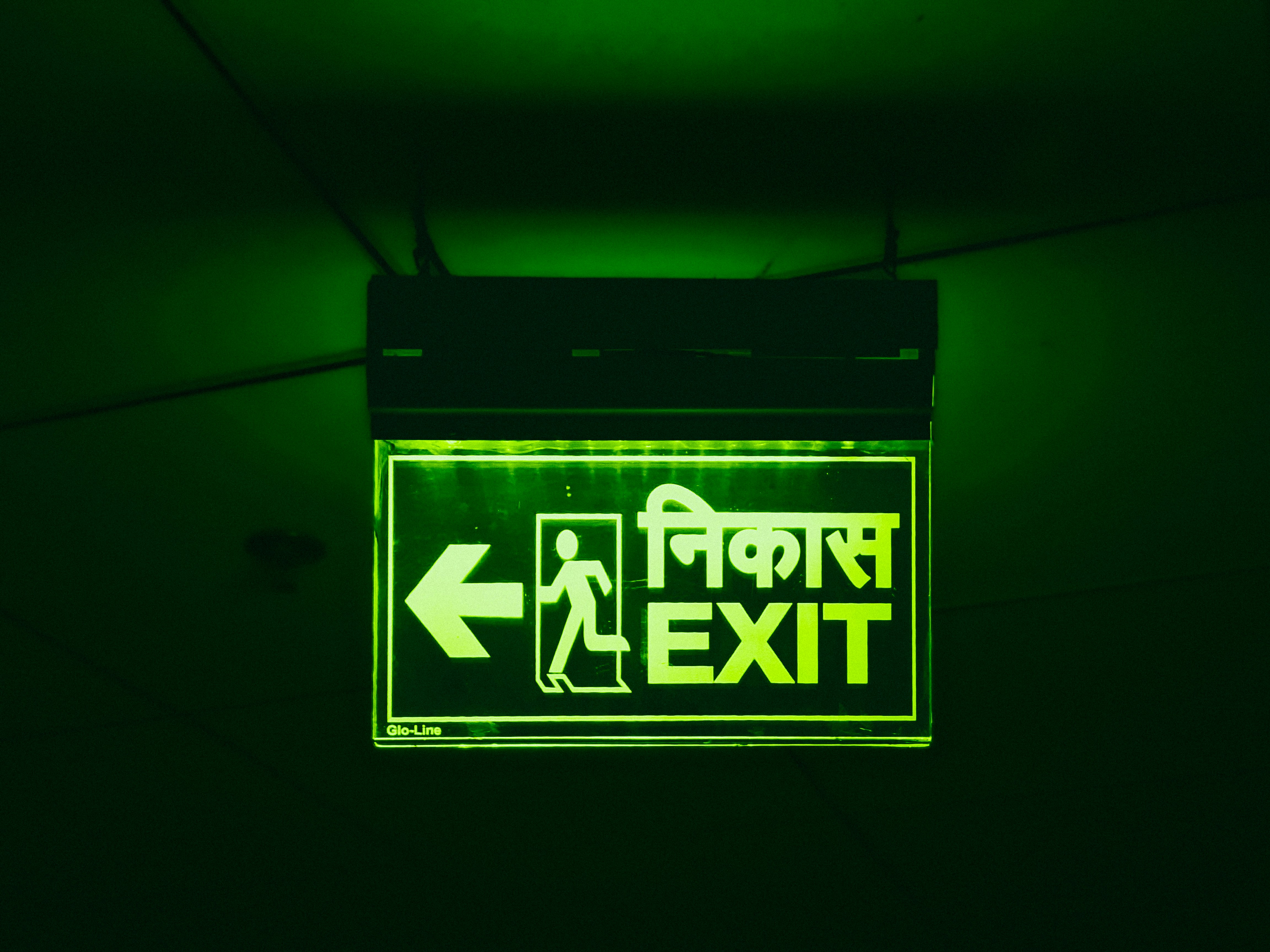 A glowing green exit sign illuminated against a dark background. | Green exit sign with arrow pointing left