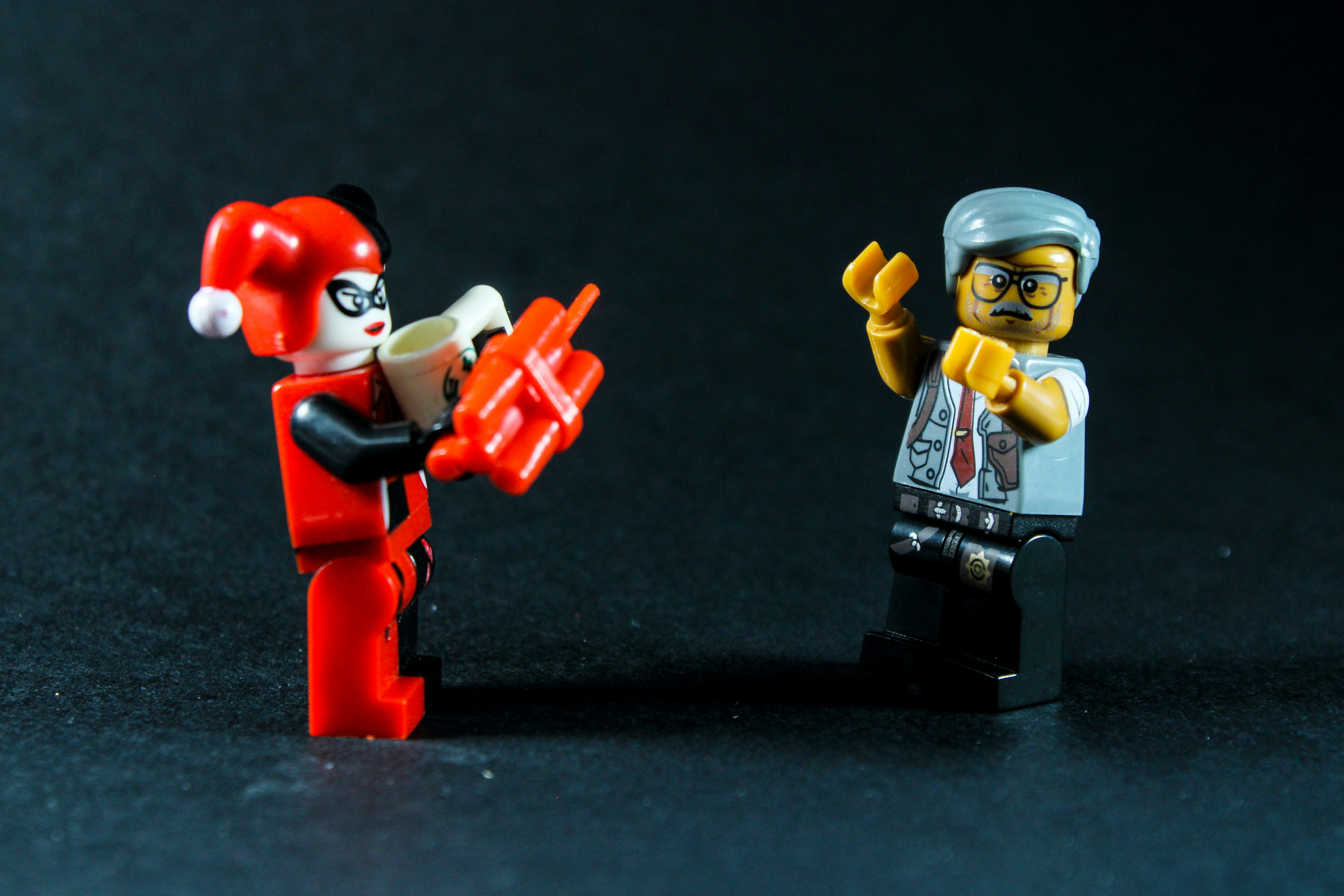 Harley Quinn and a surprised character engage in a humorous standoff with a toy weapon. The scene captures a whimsical moment between two Lego figures.