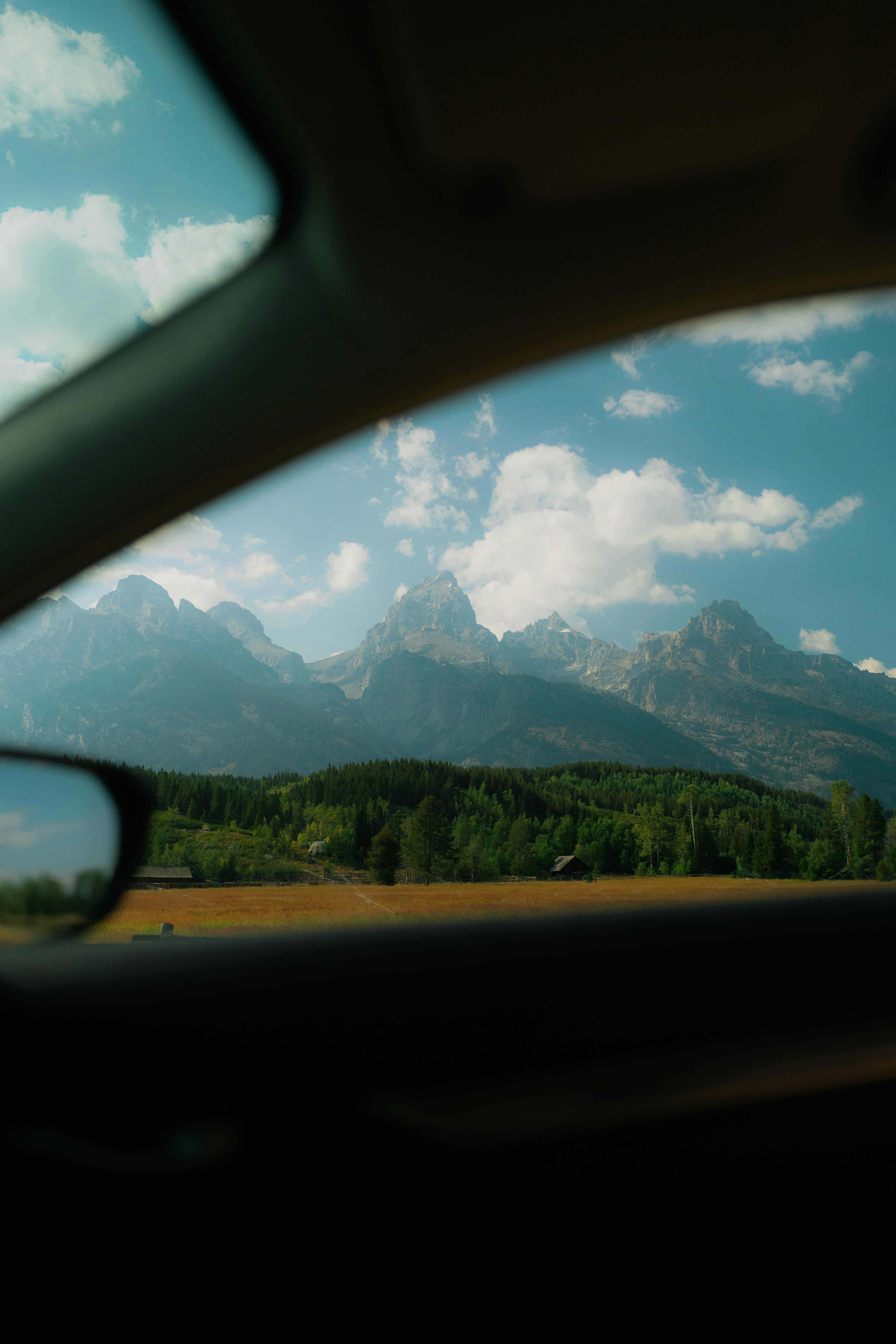 Dramatic mountain range viewed through a car window, showcasing vibrant greenery and a clear blue sky with scattered clouds.