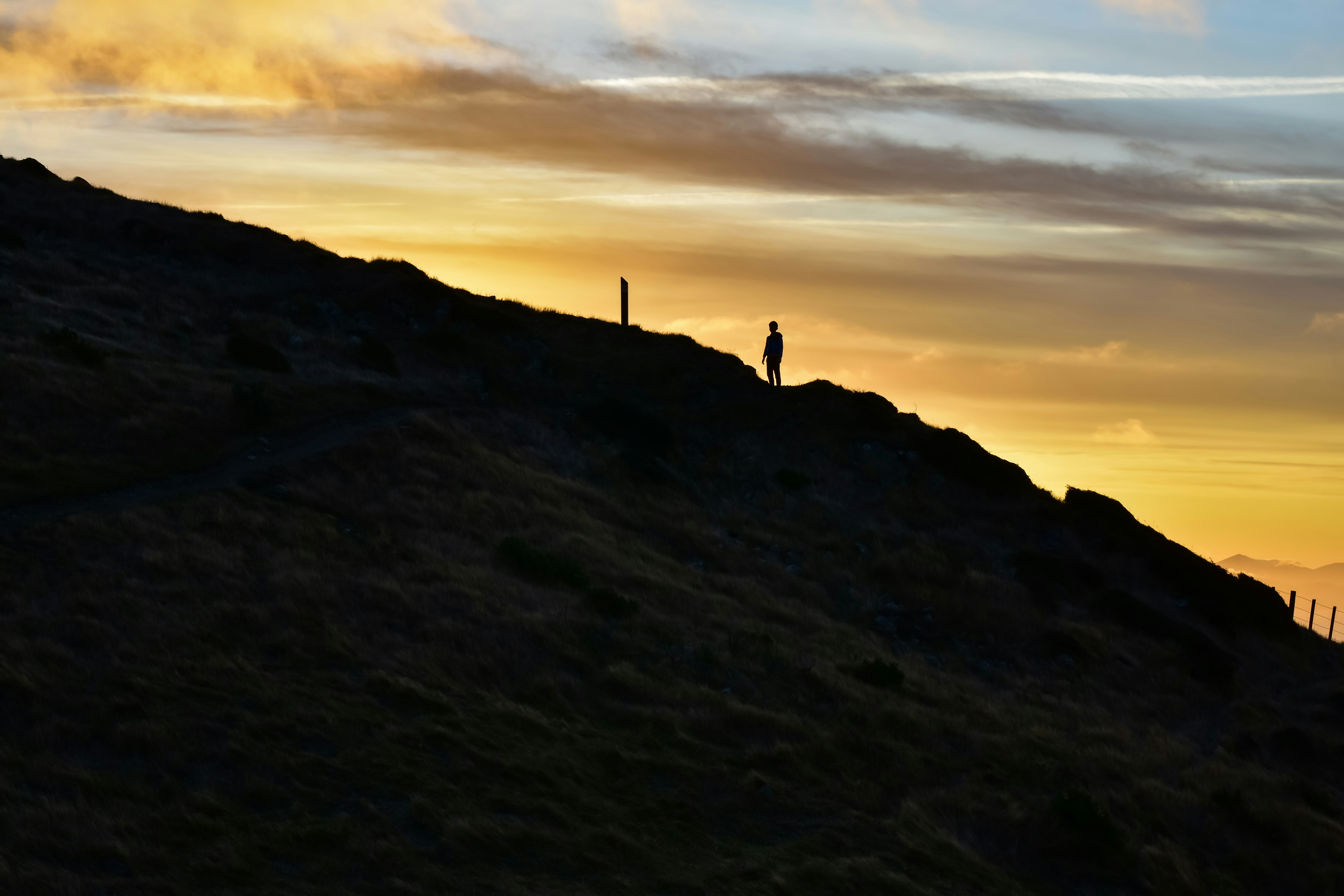 The silhouette of a single person on a hill in front of a sunset sky and clouds | Silhouette of a person walking on a grassy hill at sunset