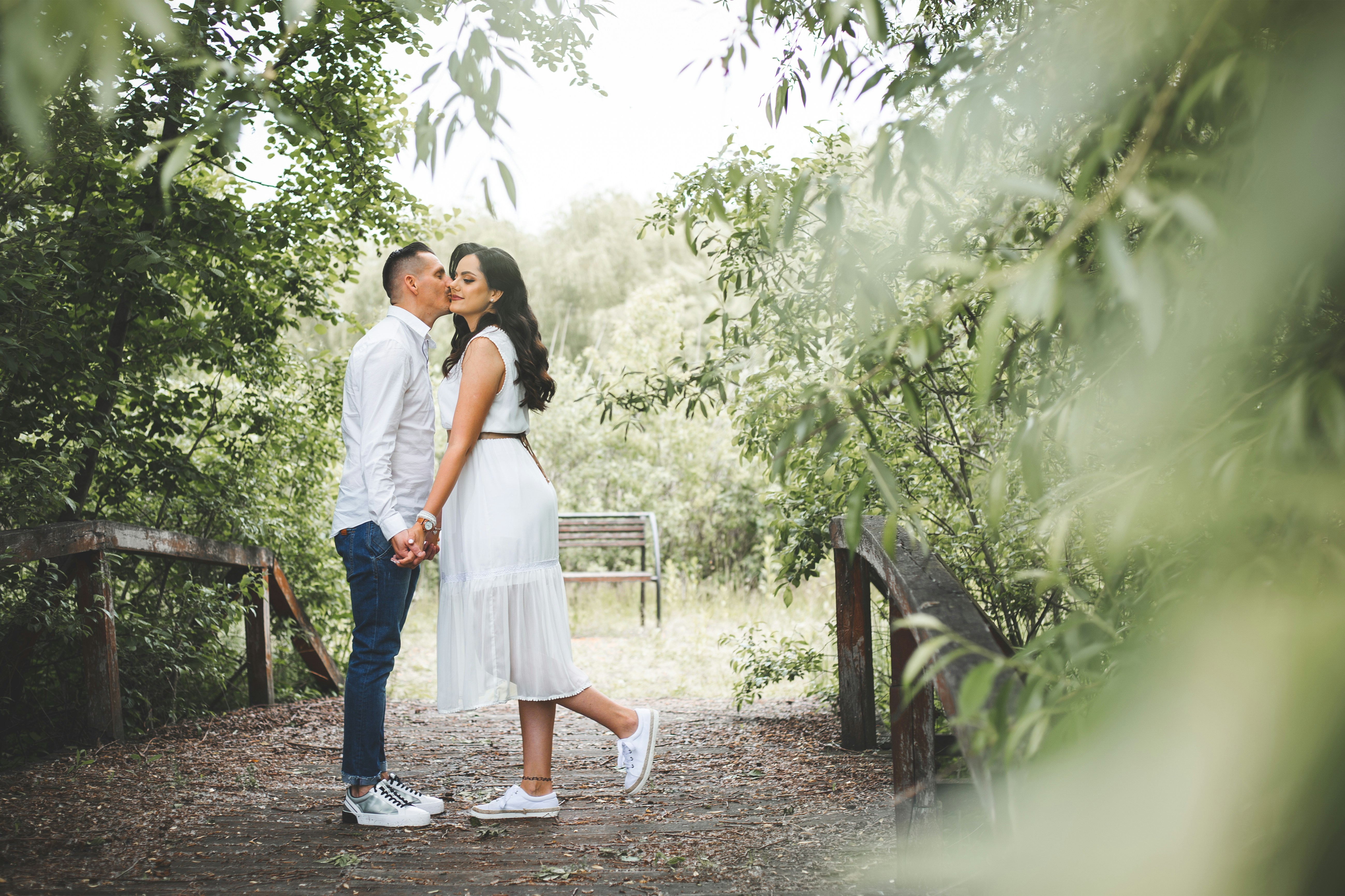Save the date photo session with the man kissing his fiancé on a bridge both holding hands. | Couple kissing on a wooden bridge surrounded by greenery