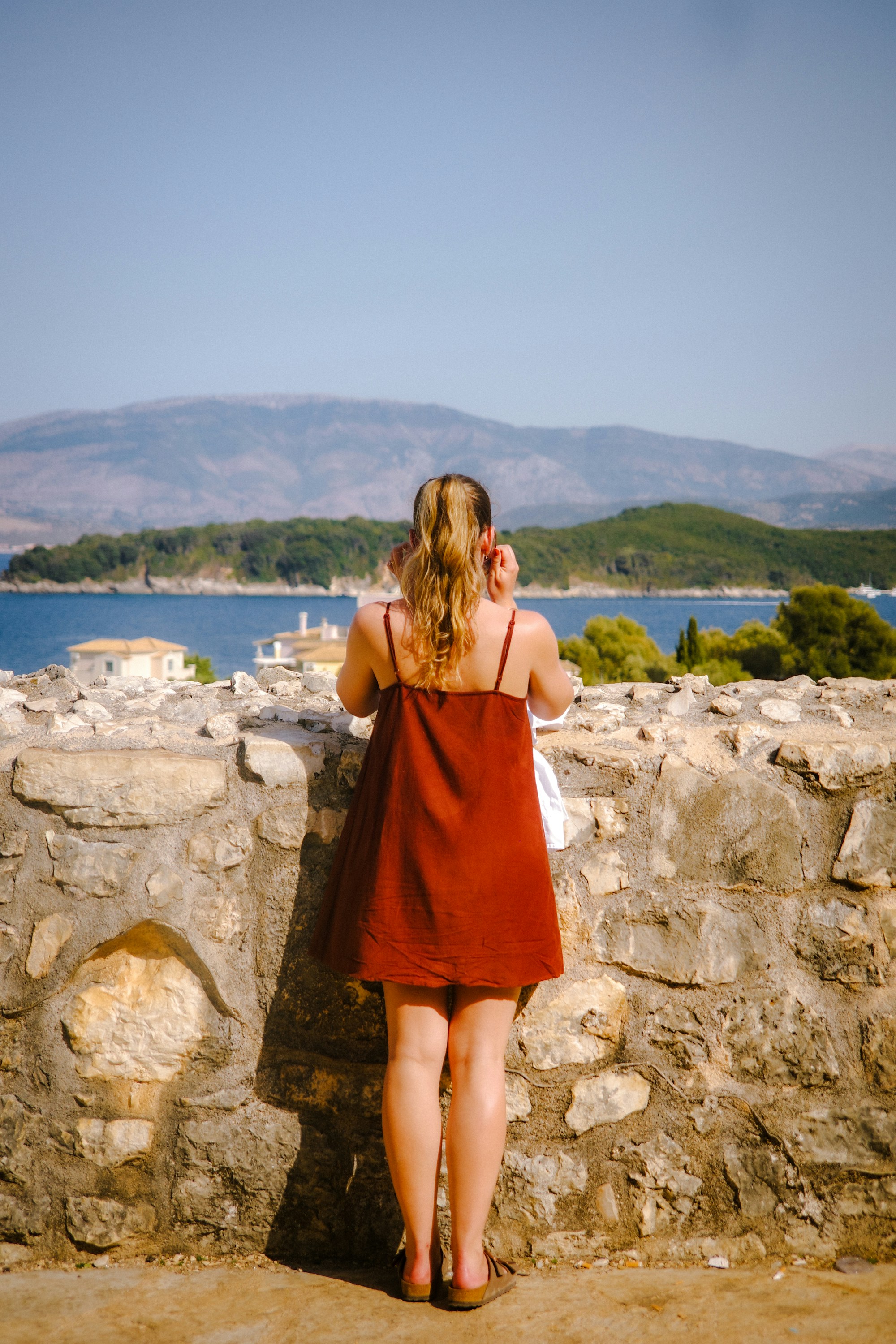 Woman in a brown dress gazing over a stone wall at a tranquil seaside view, framed by lush greenery and distant mountains.