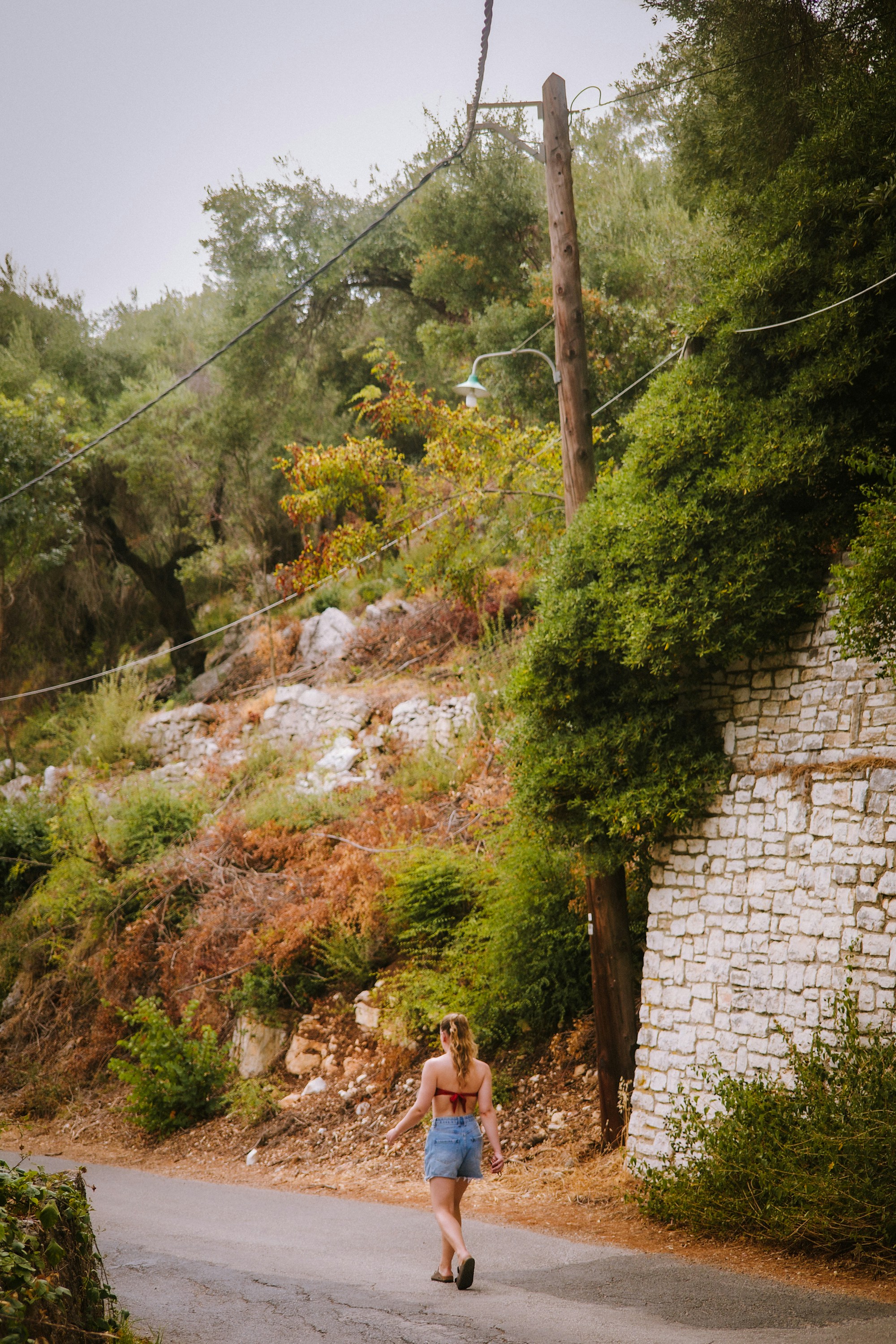 A woman strolls along a narrow road bordered by lush greenery and rustic stone walls, capturing a serene moment in a natural setting.