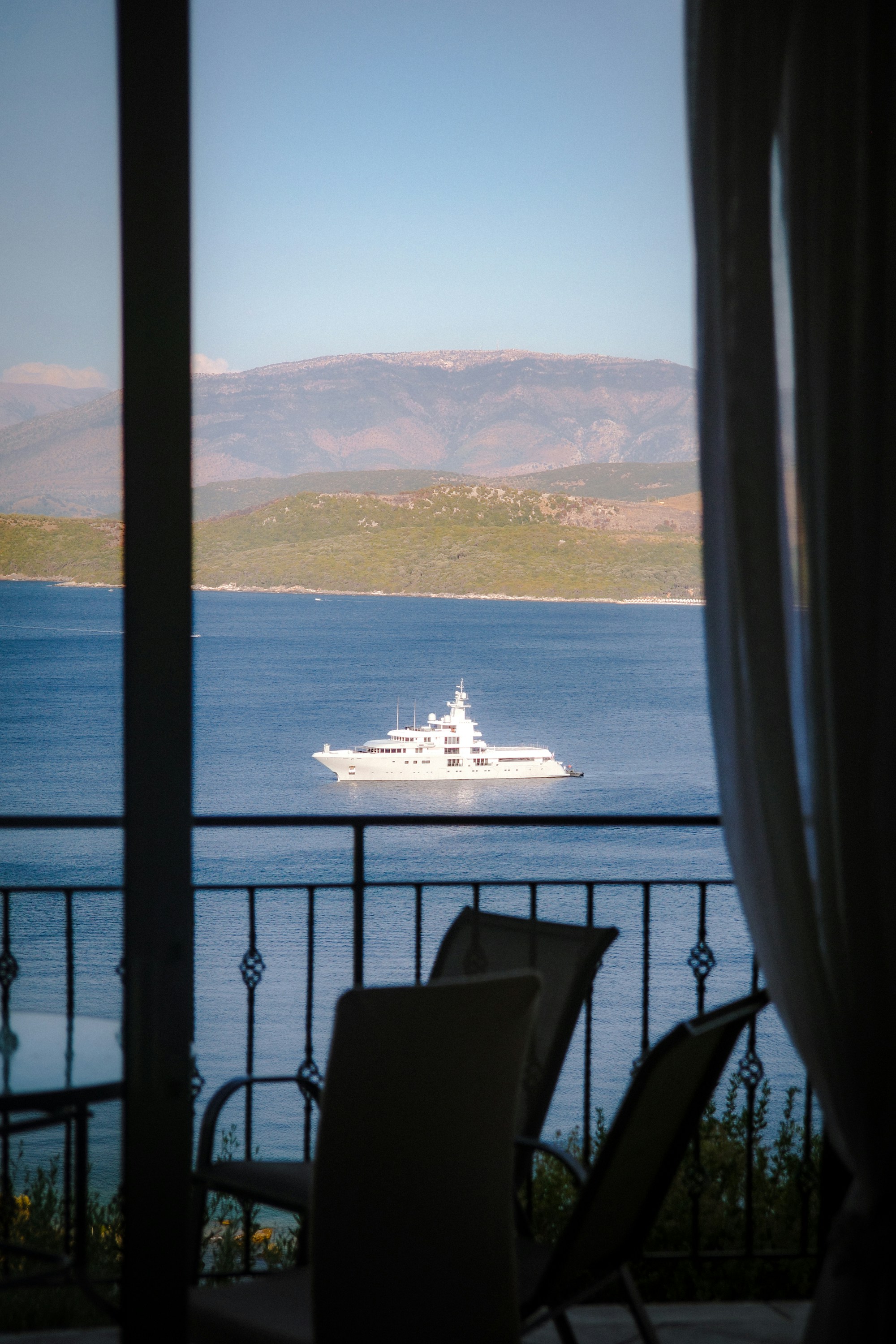 Balcony Vista: a white yacht gliding past a blue bay framed by curtains and railing