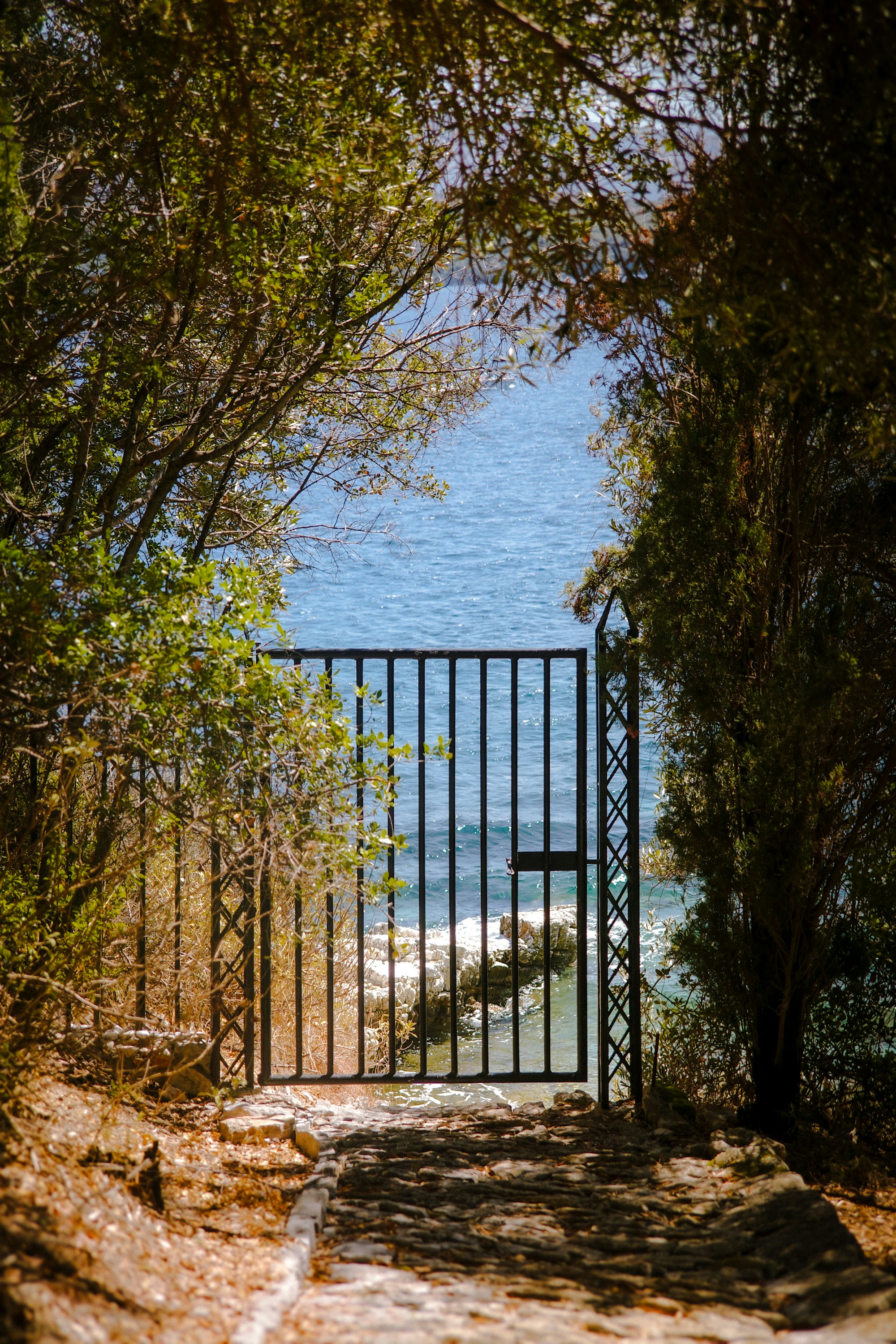 Iron gate framed by lush greenery, leading to a serene coastal view. The calm waters glisten in the background, inviting exploration.