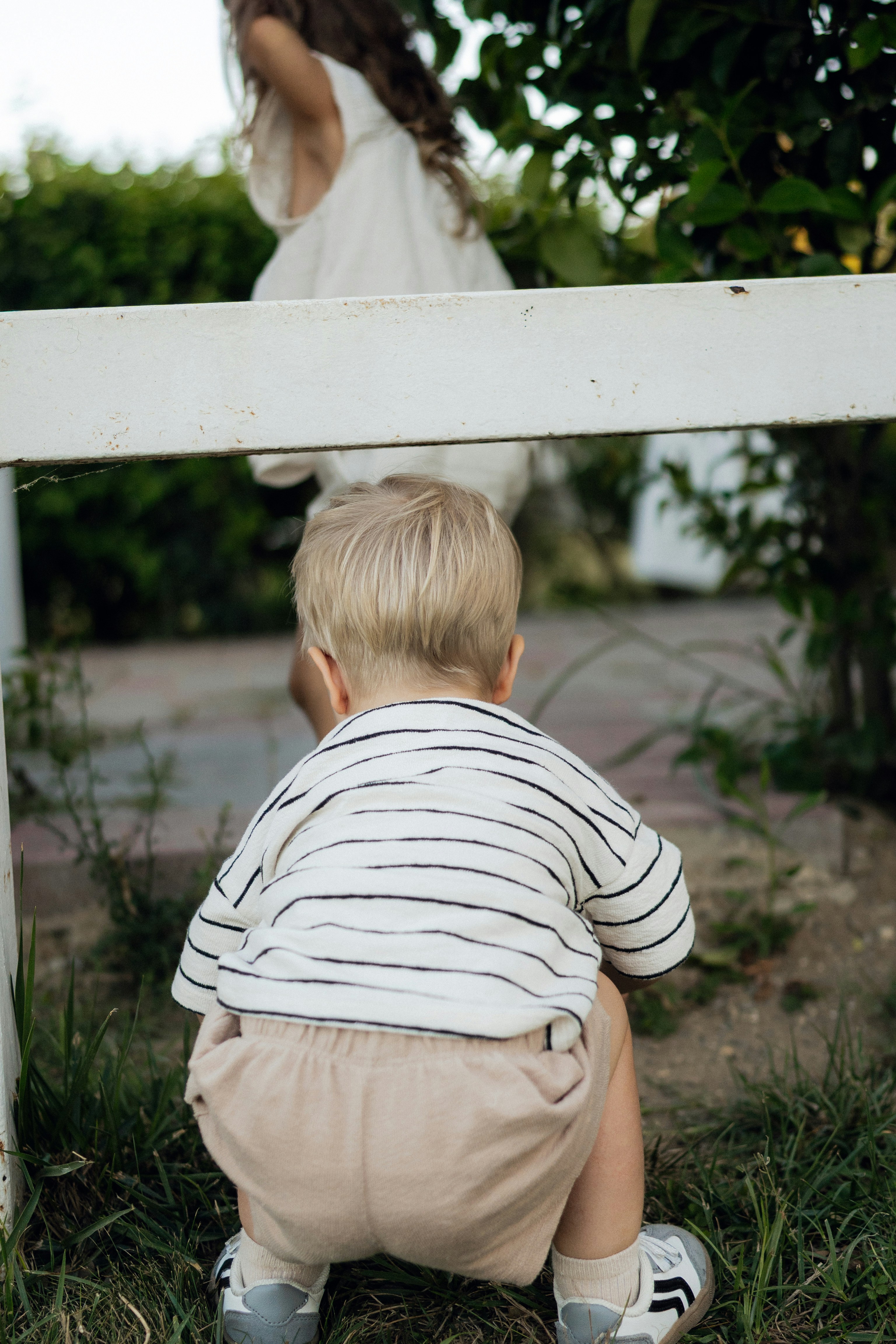 Young child crouching near a garden gate while a figure in a white dress moves away, creating a sense of adventure and curiosity.