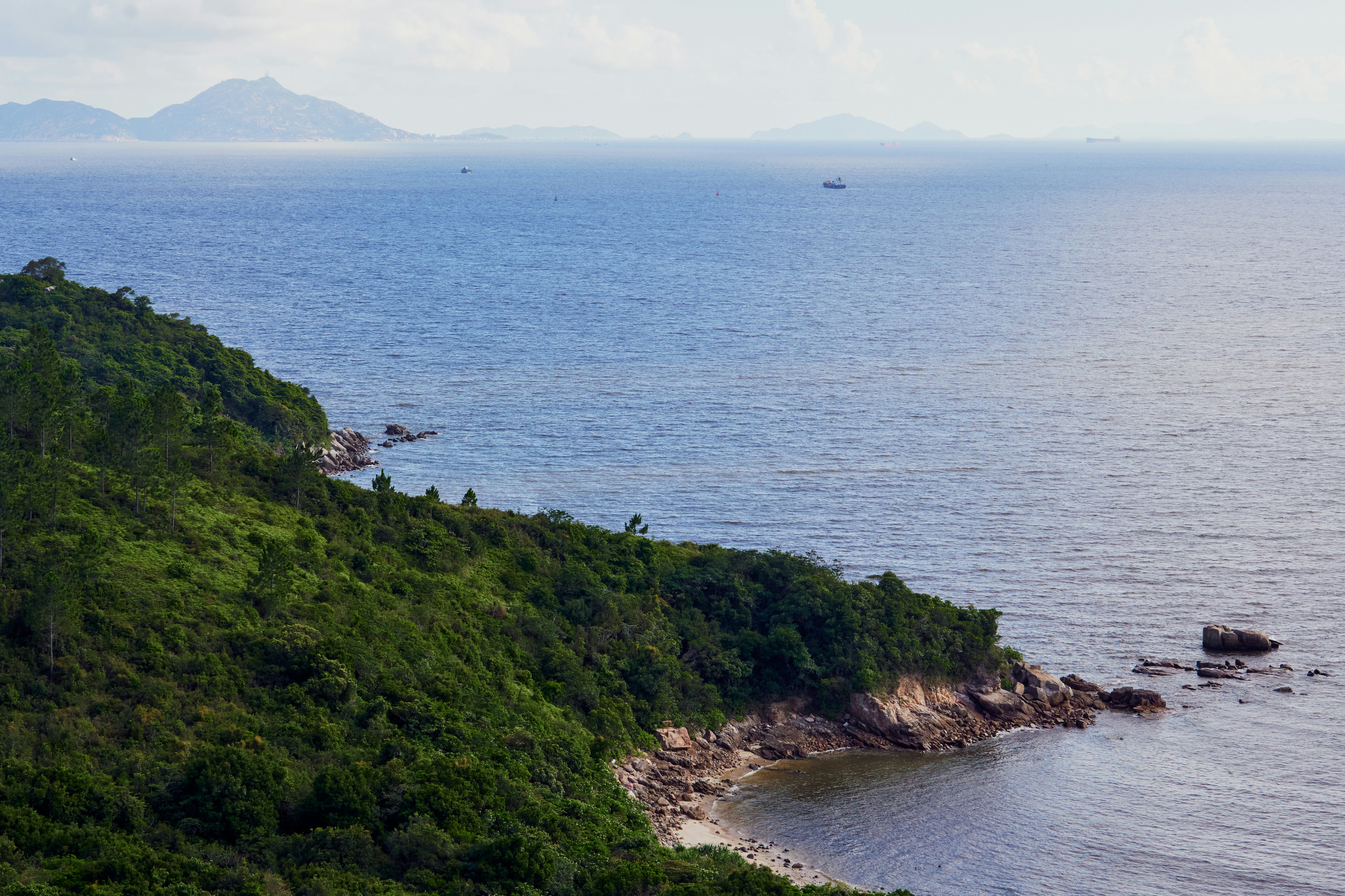 Lush green coastline meeting the tranquil sea under a bright sky, with distant mountains on the horizon. A few boats dot the water's surface.