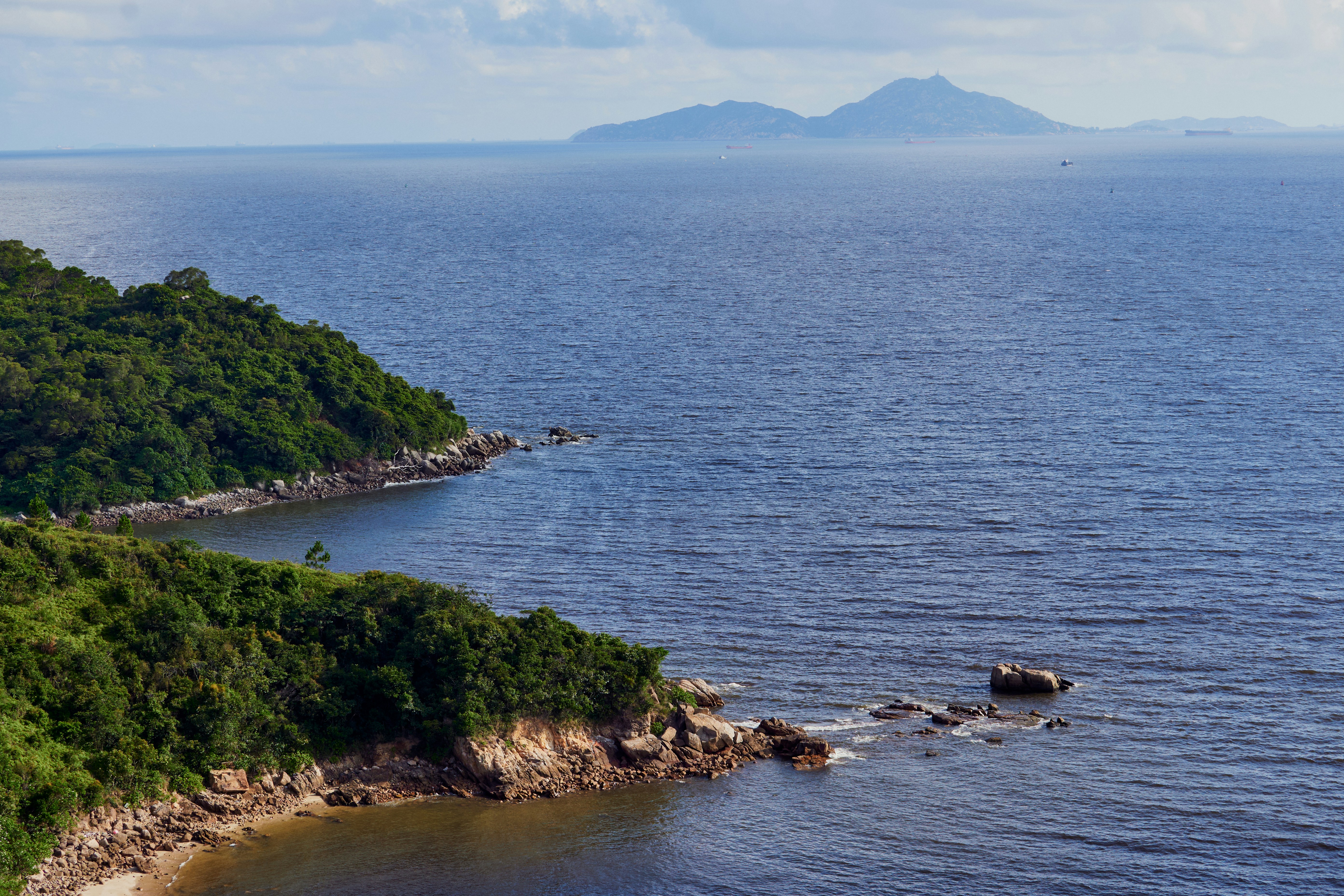 Lush green coastline meets the tranquil sea under a bright sky, with distant mountains visible on the horizon.